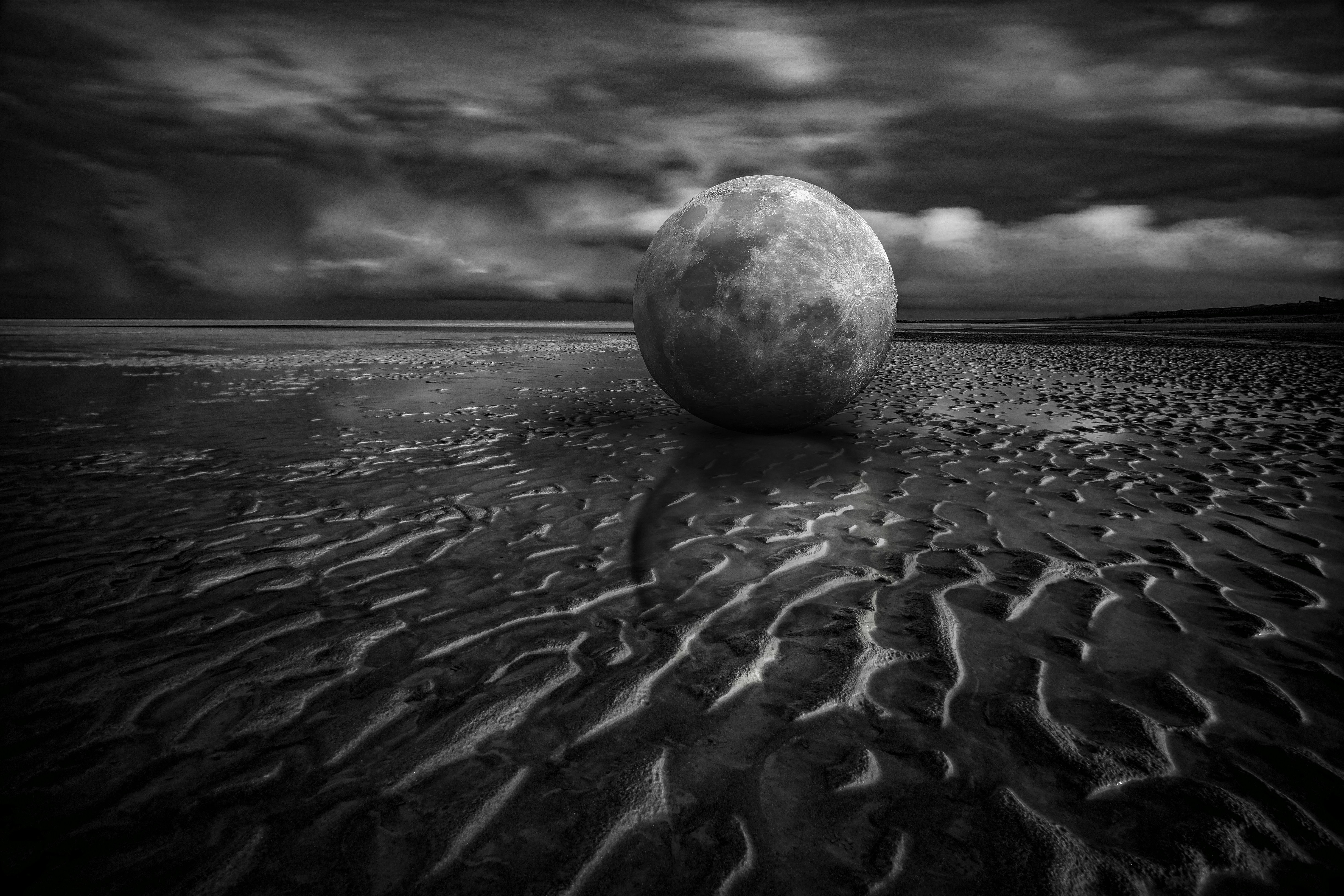 This surreal black-and-white image captures a giant moon resting on rippled tidal flats, creating an otherworldly atmosphere. The textured foreground contrasts with the smooth celestial sphere, under a dramatic cloudy sky. The monochrome palette enhances the mystical mood, highlighting the intricate patterns and shadows that make this scene visually captivating.