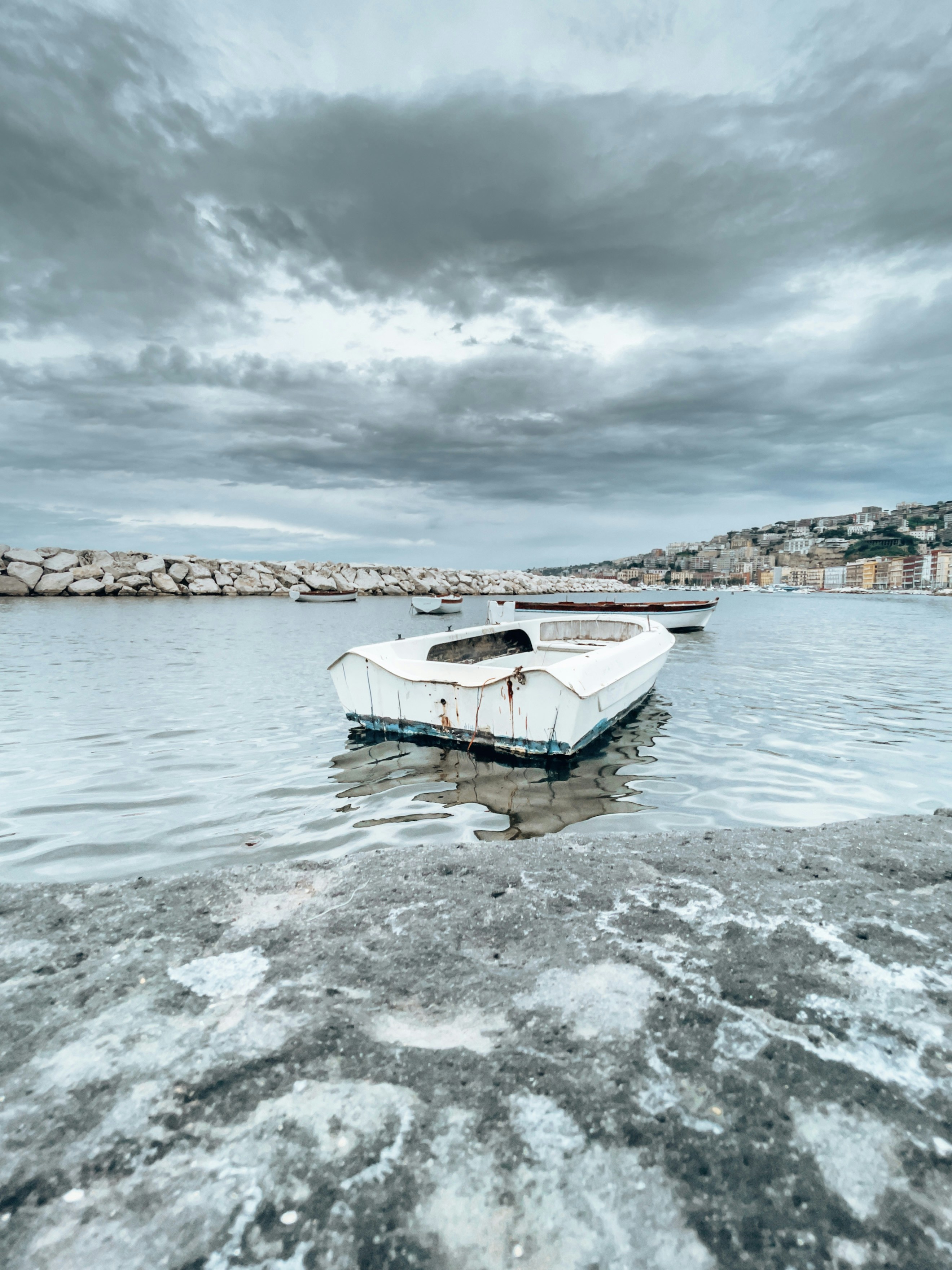 Weathered boat floating in calm waters with a backdrop of a coastal town and dramatic clouds. The scene captures a moment of tranquility amidst urban life.