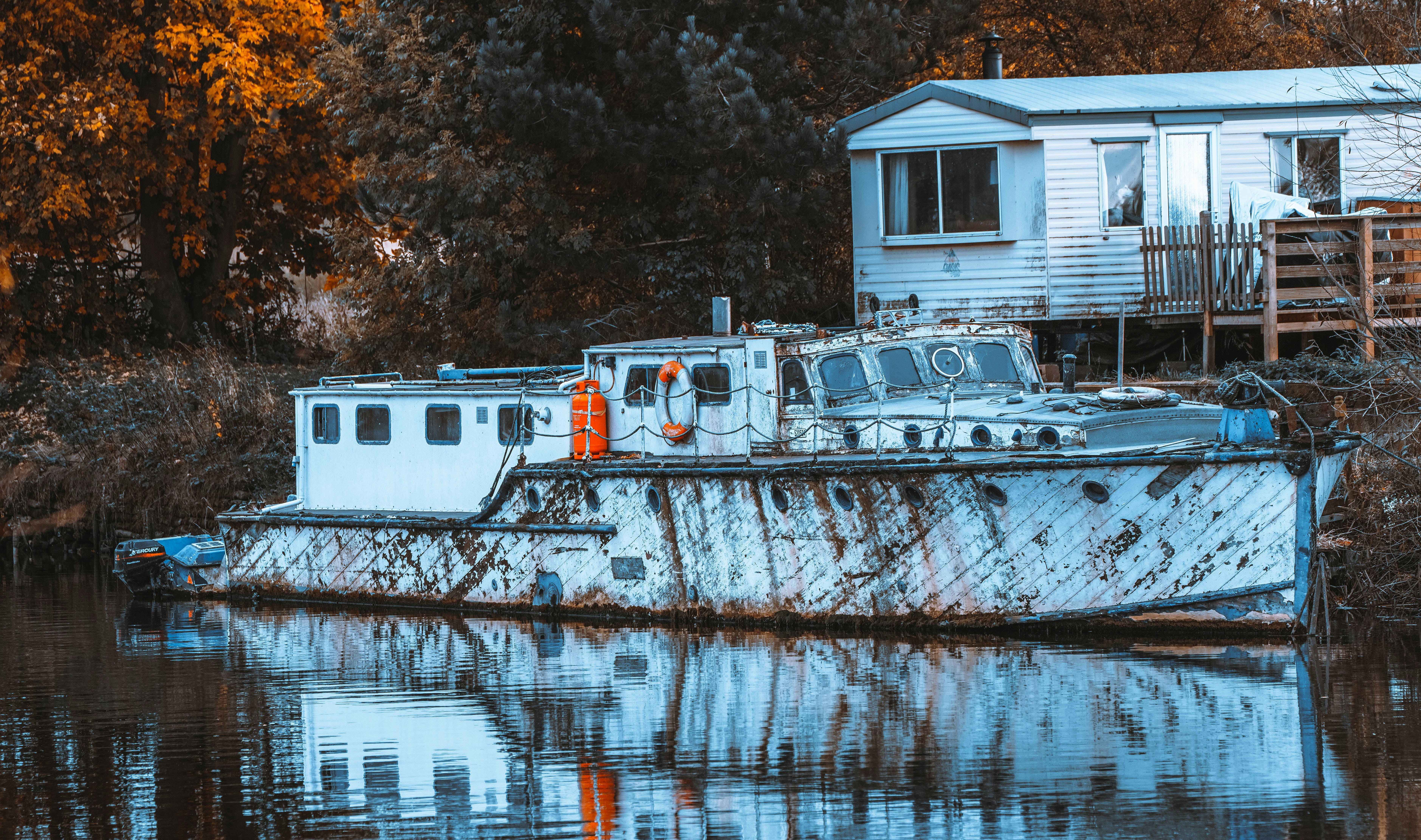 An abandoned boat rests quietly on a still river, surrounded by autumn foliage and a nearby cabin. The scene captures the interplay of rust and reflection in tranquil waters.