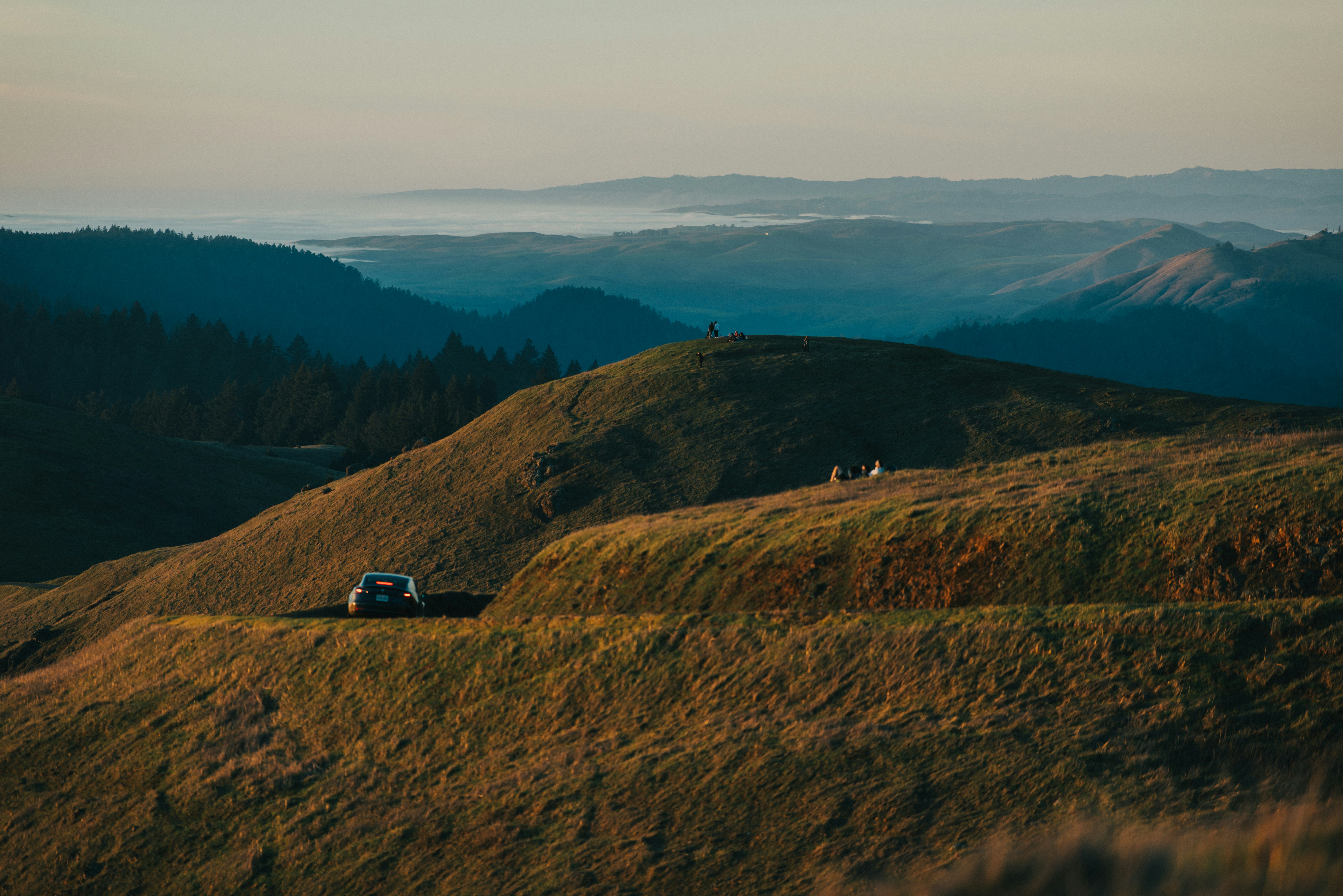 A vehicle navigates a winding road atop grassy hills, with distant mountains fading into the horizon. The soft evening light enhances the serene landscape.