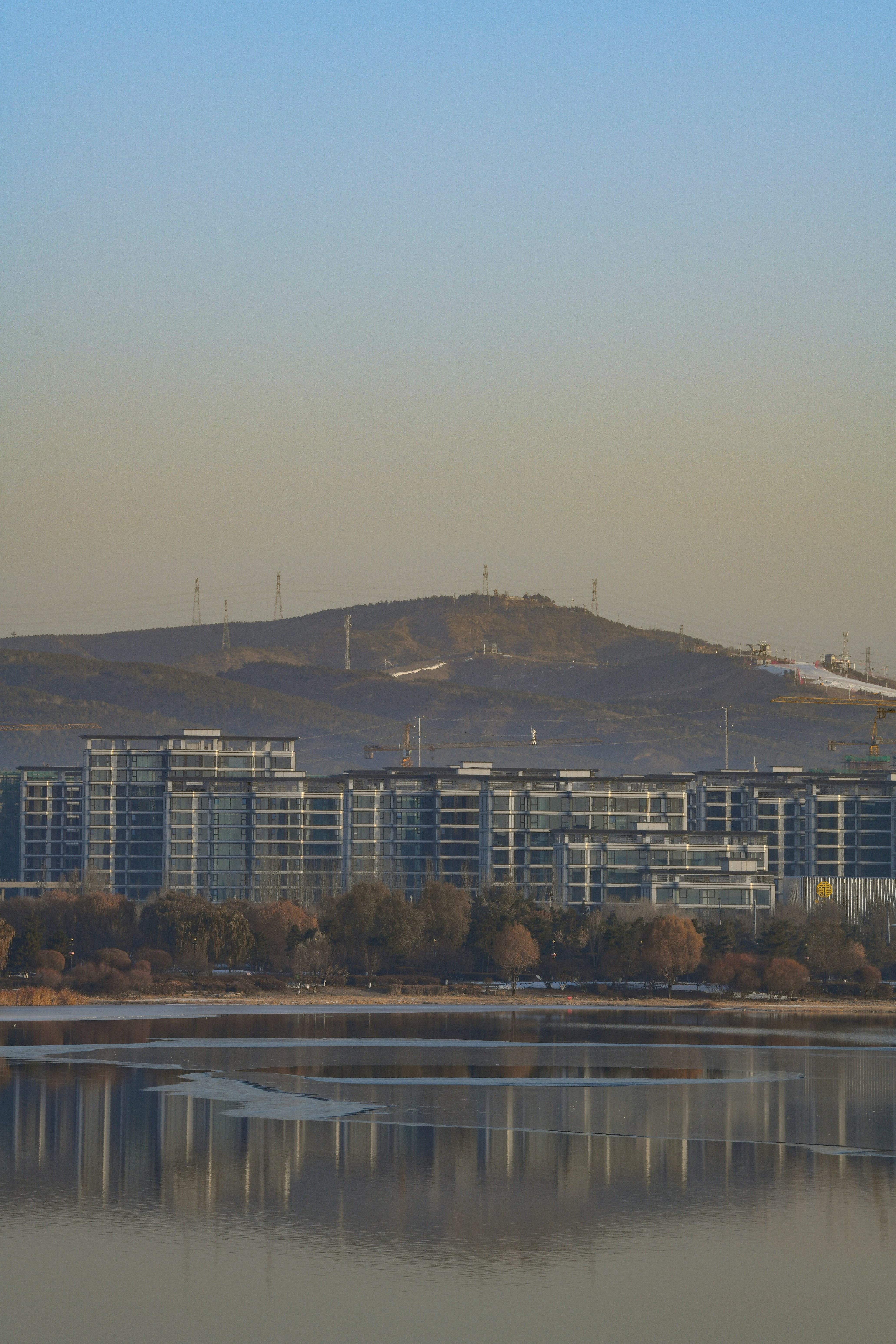 Modern residential buildings reflecting on a calm lake with distant hills in the background.