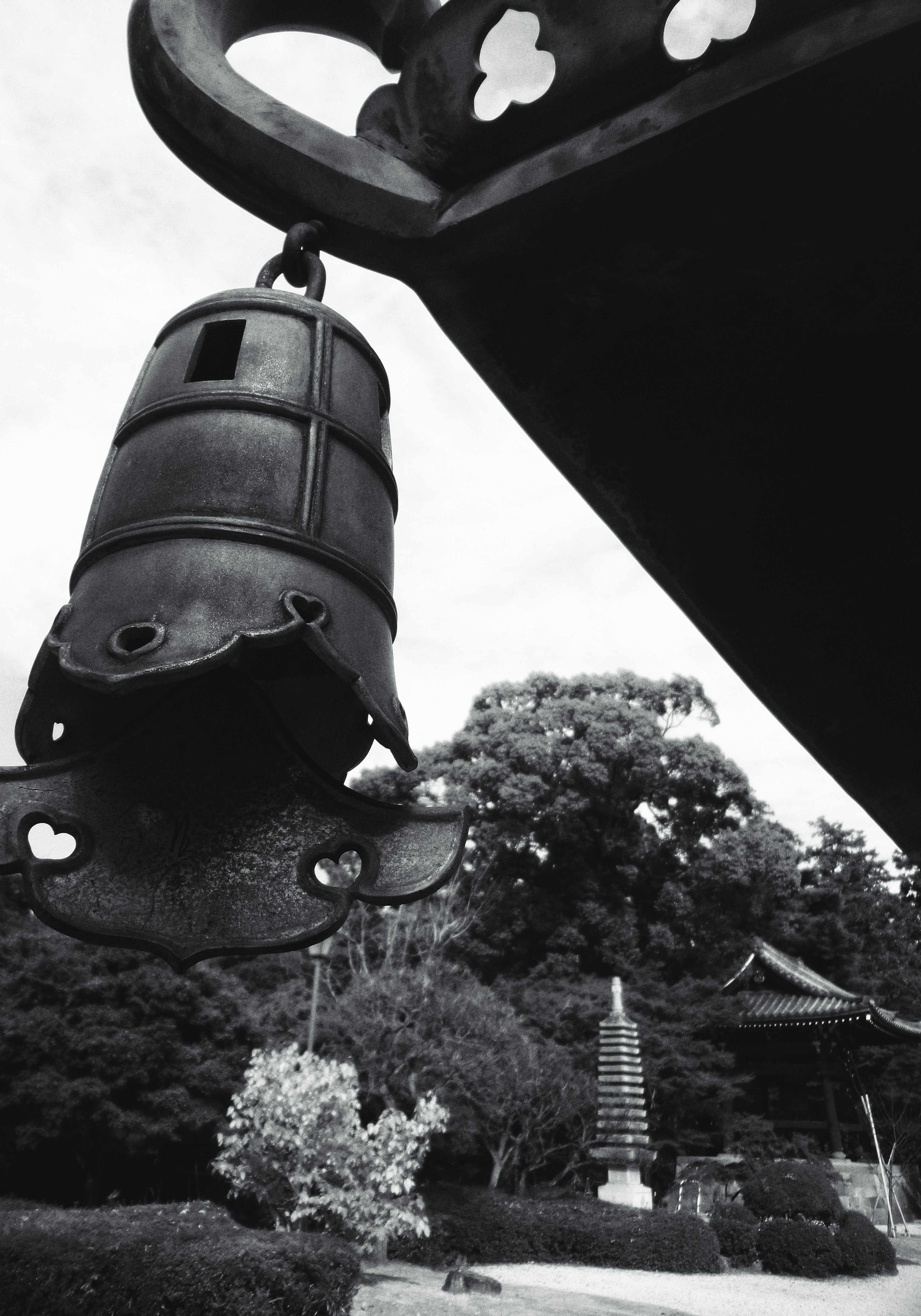 Intricately designed bell suspended under a traditional structure, surrounded by lush greenery and historical elements in the background.