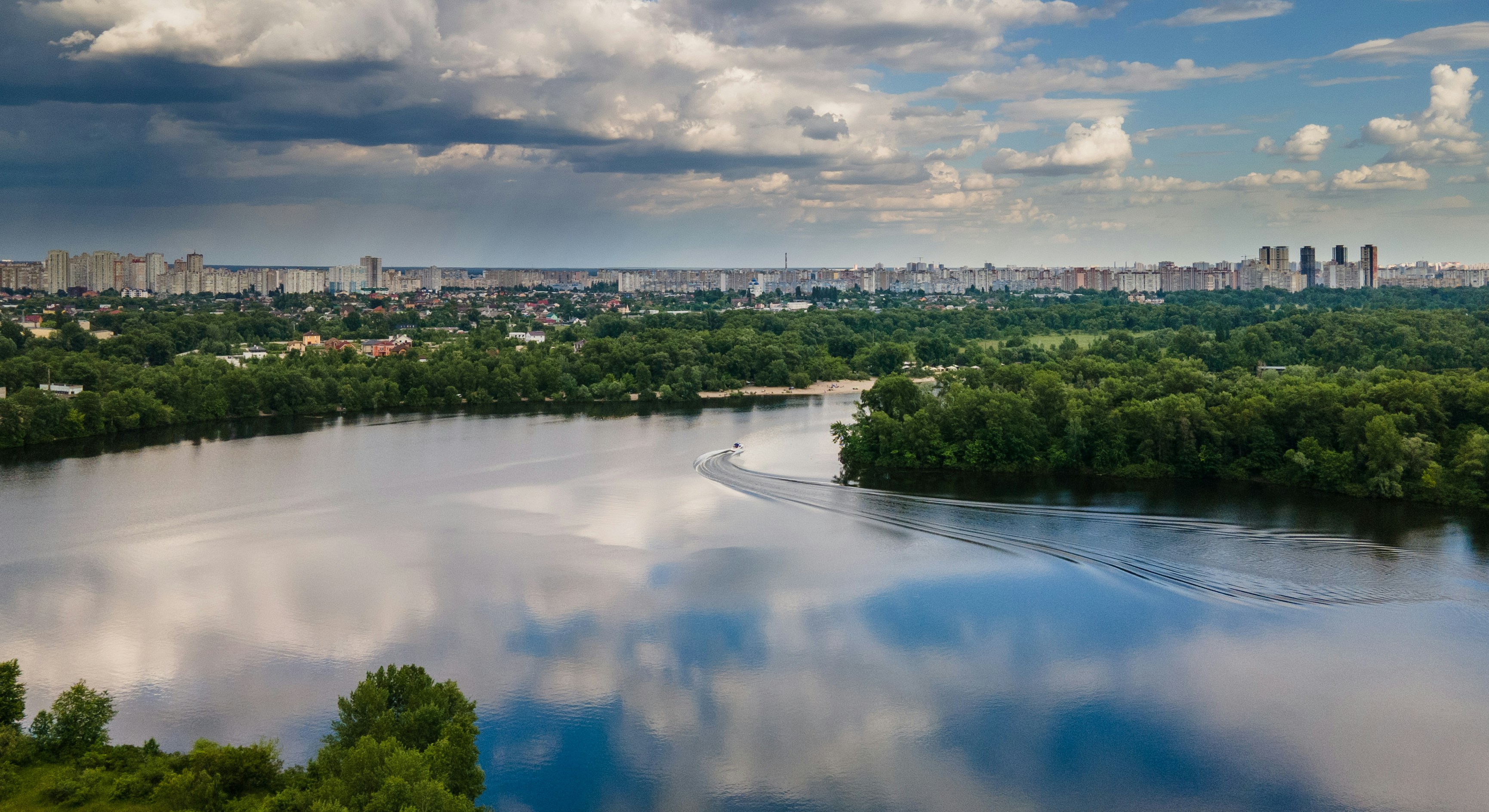 A tranquil river meandering through lush greenery, reflecting the clouds and city skyline in the background.
