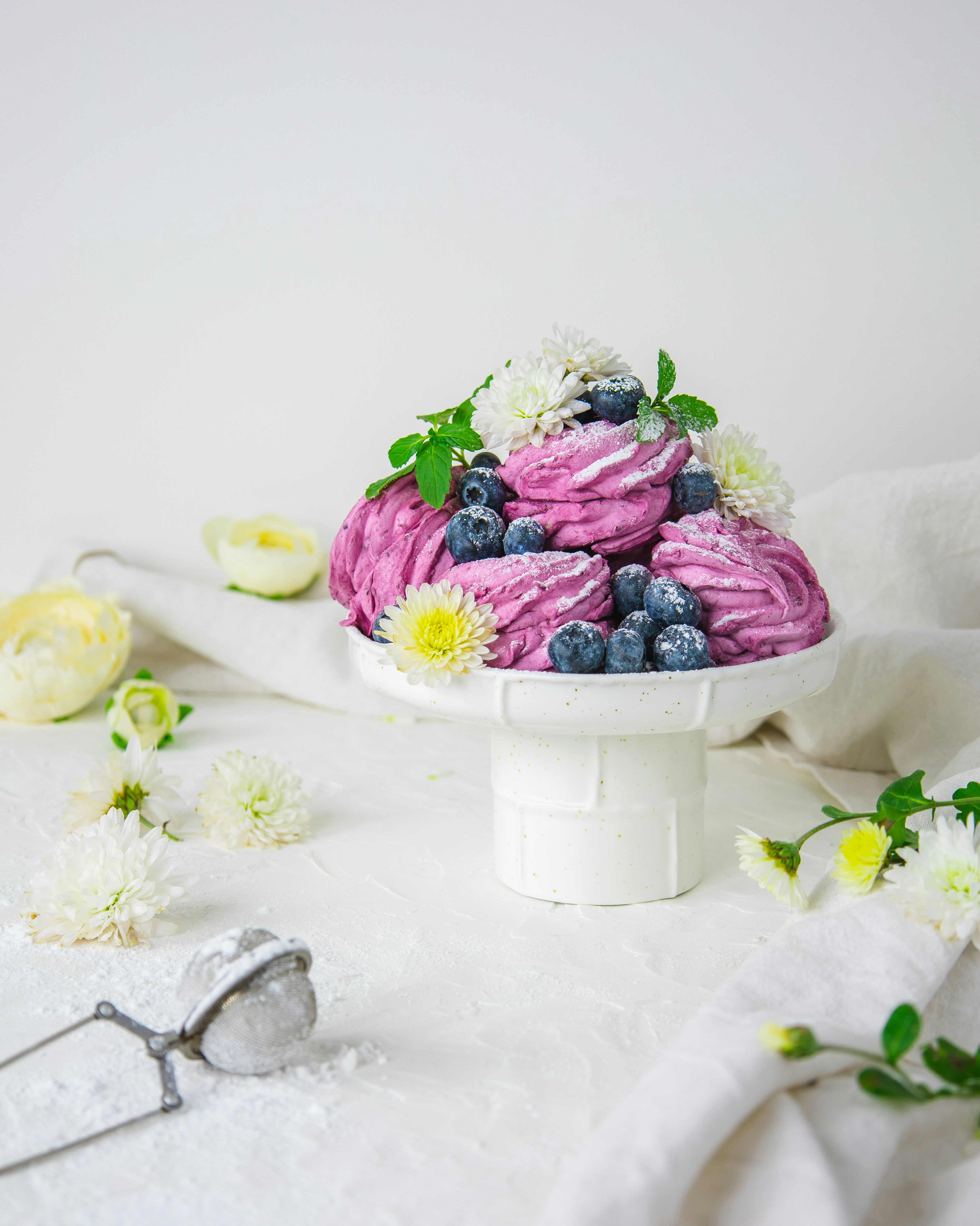 A vibrant dessert featuring pink meringues topped with blueberries and delicate flowers, elegantly presented on a white pedestal. The soft textures and colors create a visually appealing composition.