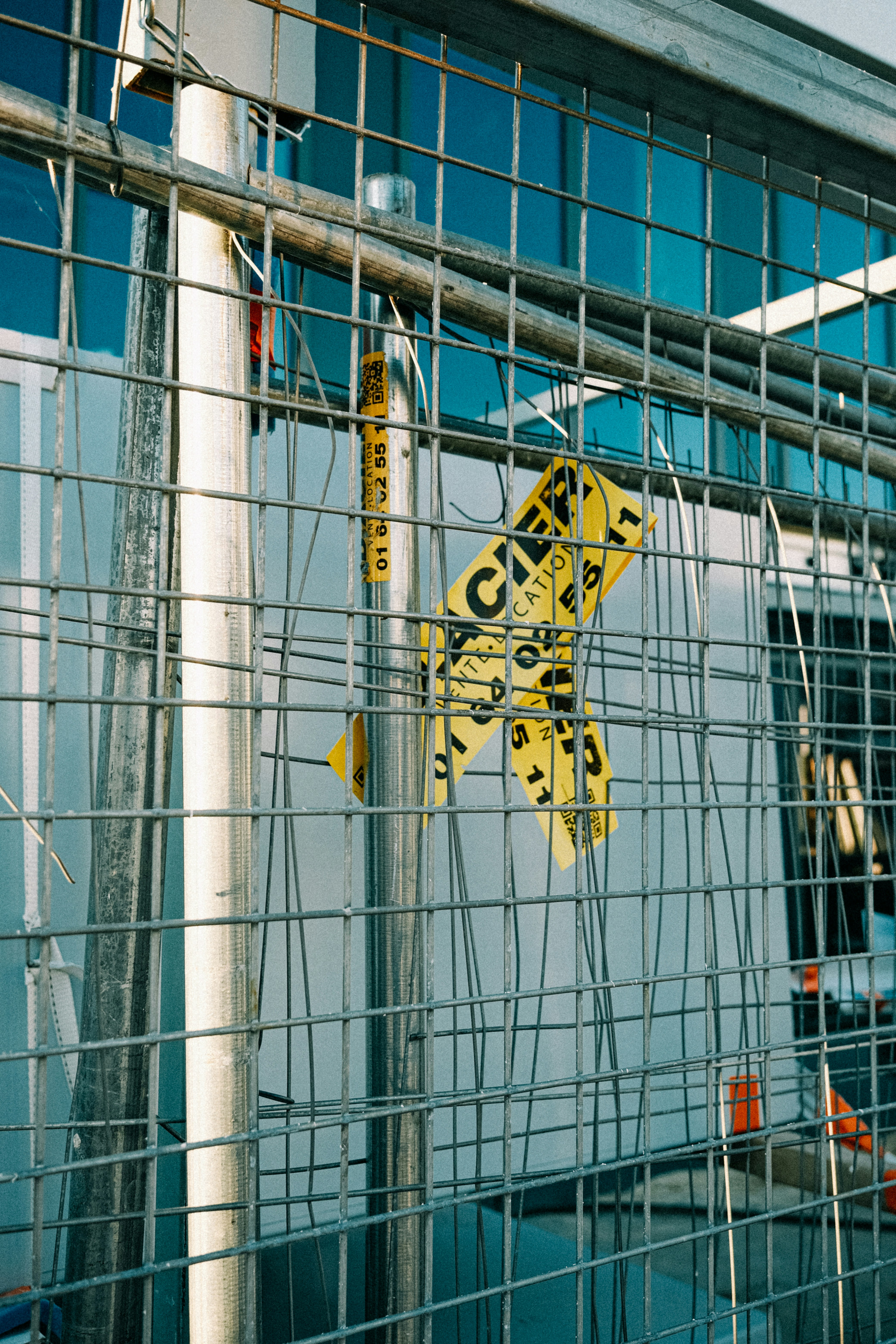 Yellow caution signs affixed to a metal fence, juxtaposed against a modern building backdrop.
