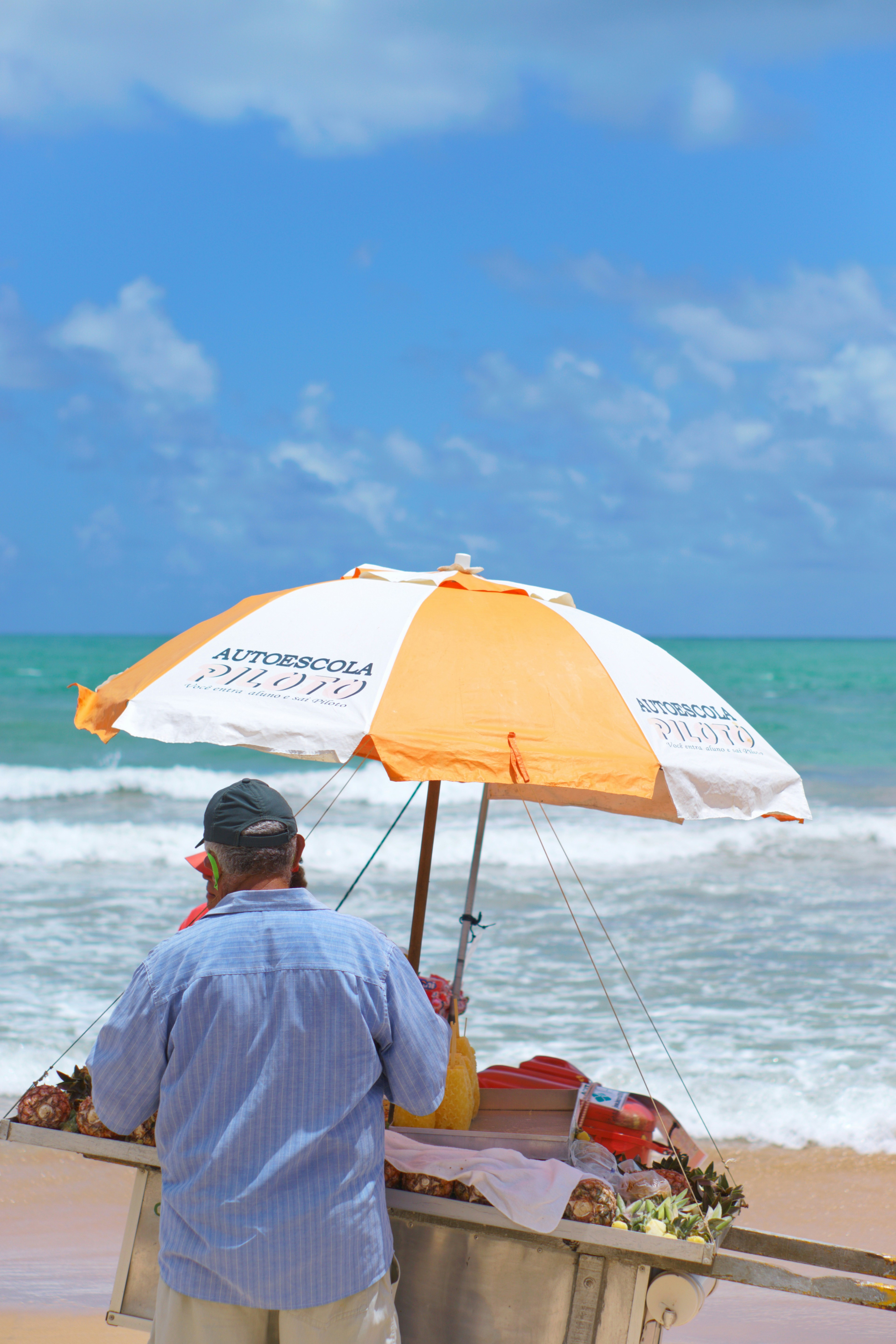 Vendor under a vibrant orange and white umbrella selling fresh fruits on a sunny beach. Gentle waves lap at the shore in the background.