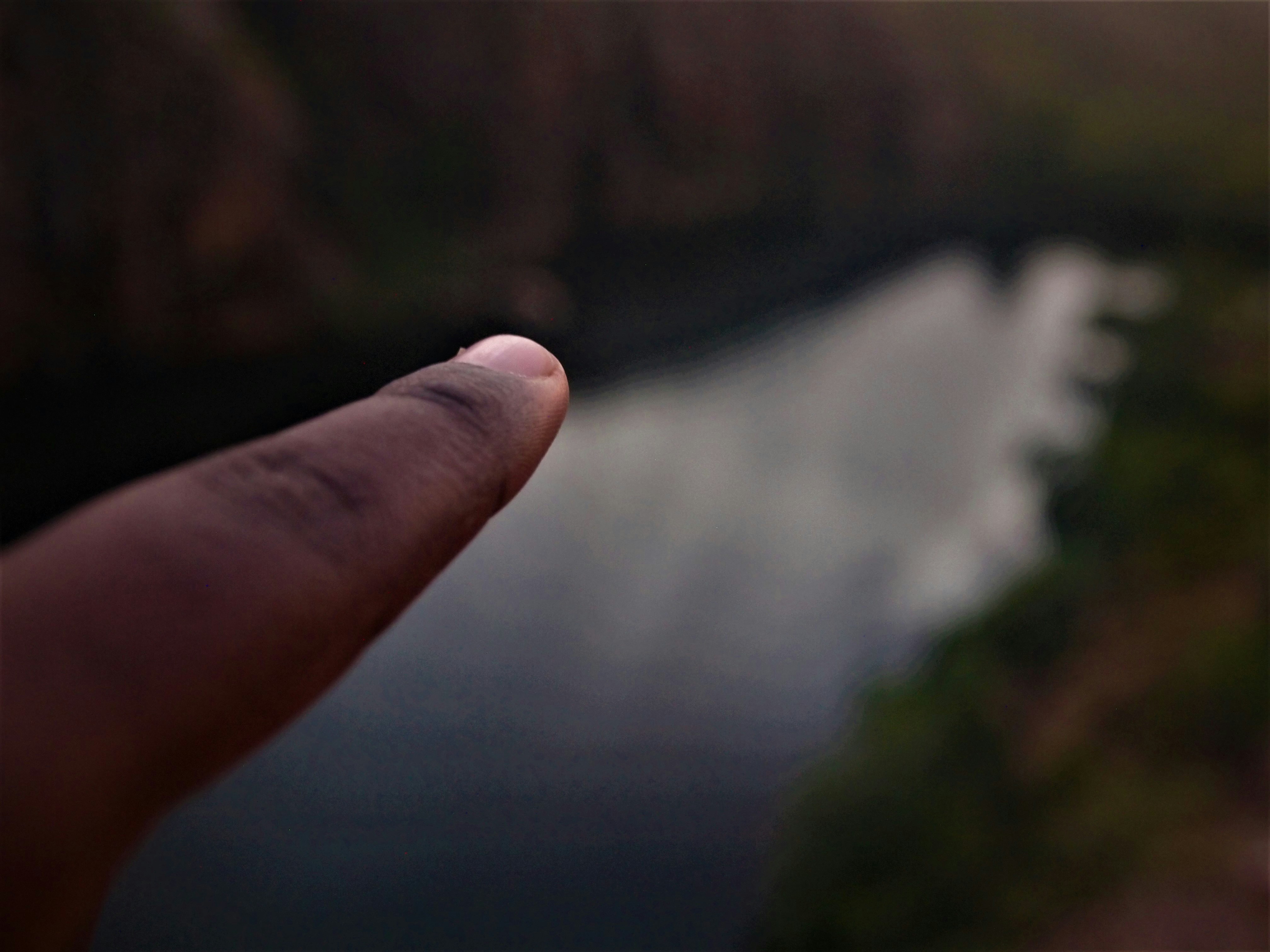 A finger points towards a serene body of water reflecting soft hues, surrounded by lush greenery and rocky terrain.