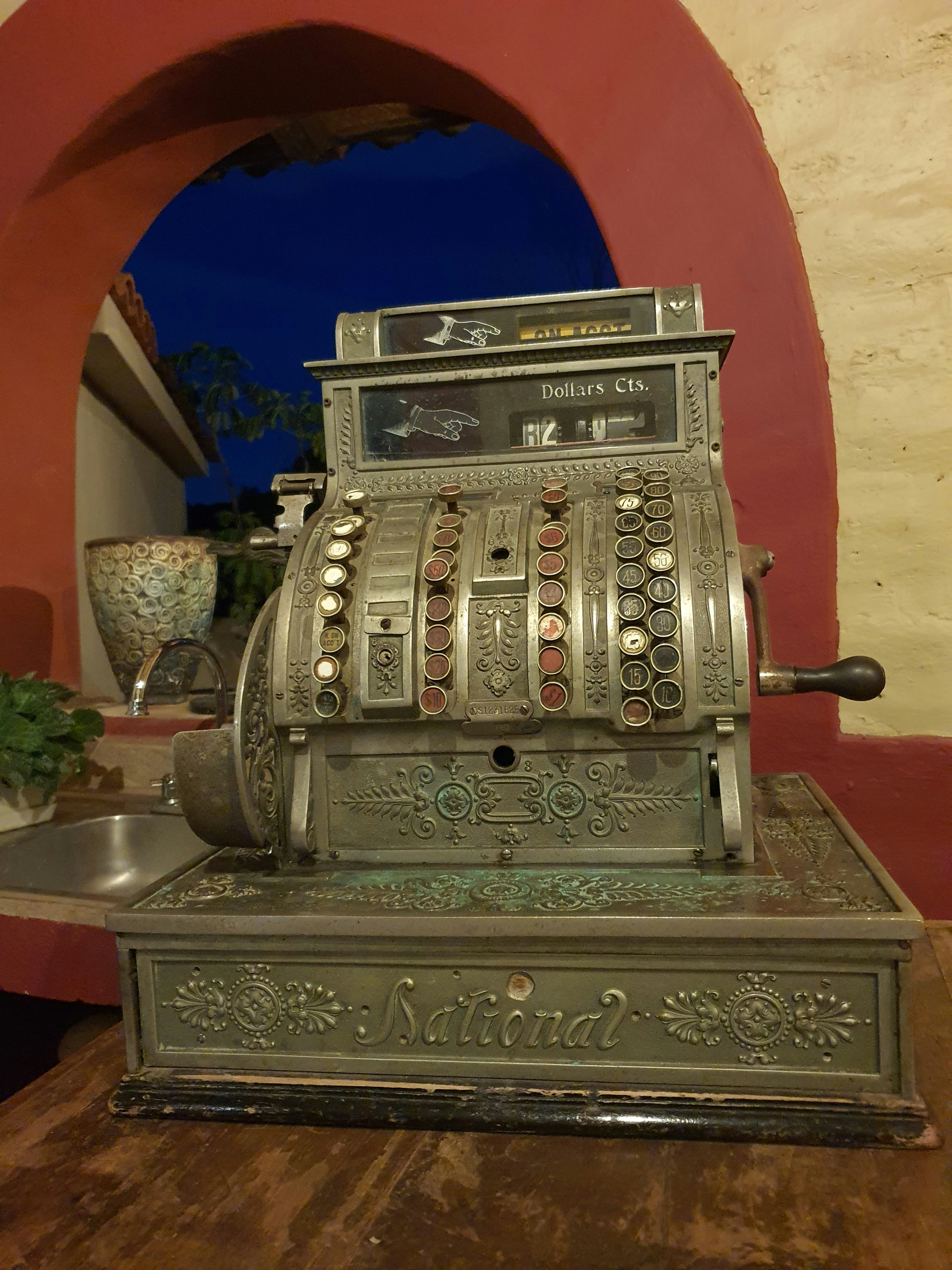 Vintage cash register with intricate detailing and colorful buttons, set against a rustic background. The warm lighting highlights its aged metal finish.
