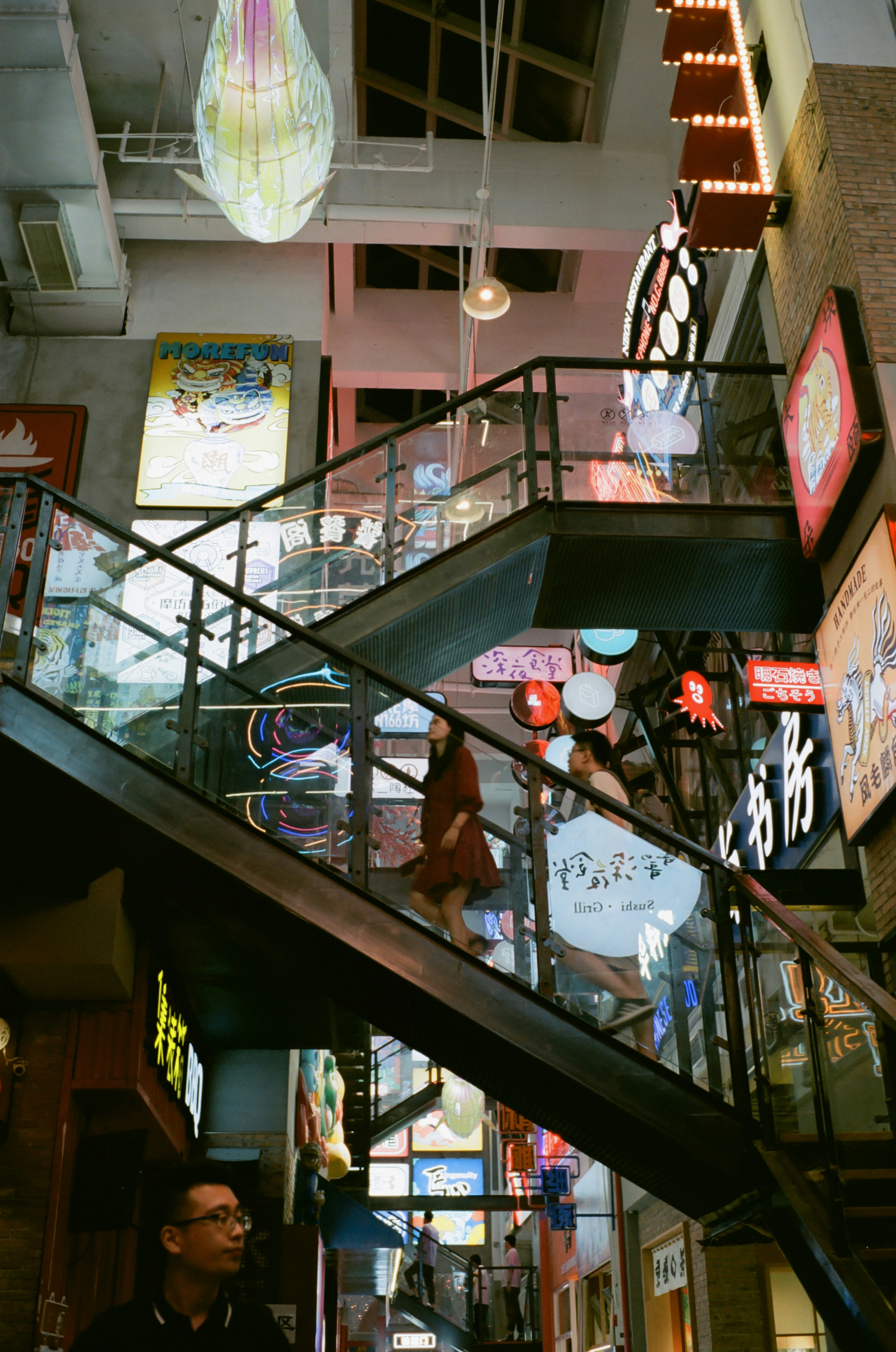 Two figures ascend a staircase lined with vibrant neon signs in a bustling urban setting. The scene captures the lively atmosphere of a modern marketplace.