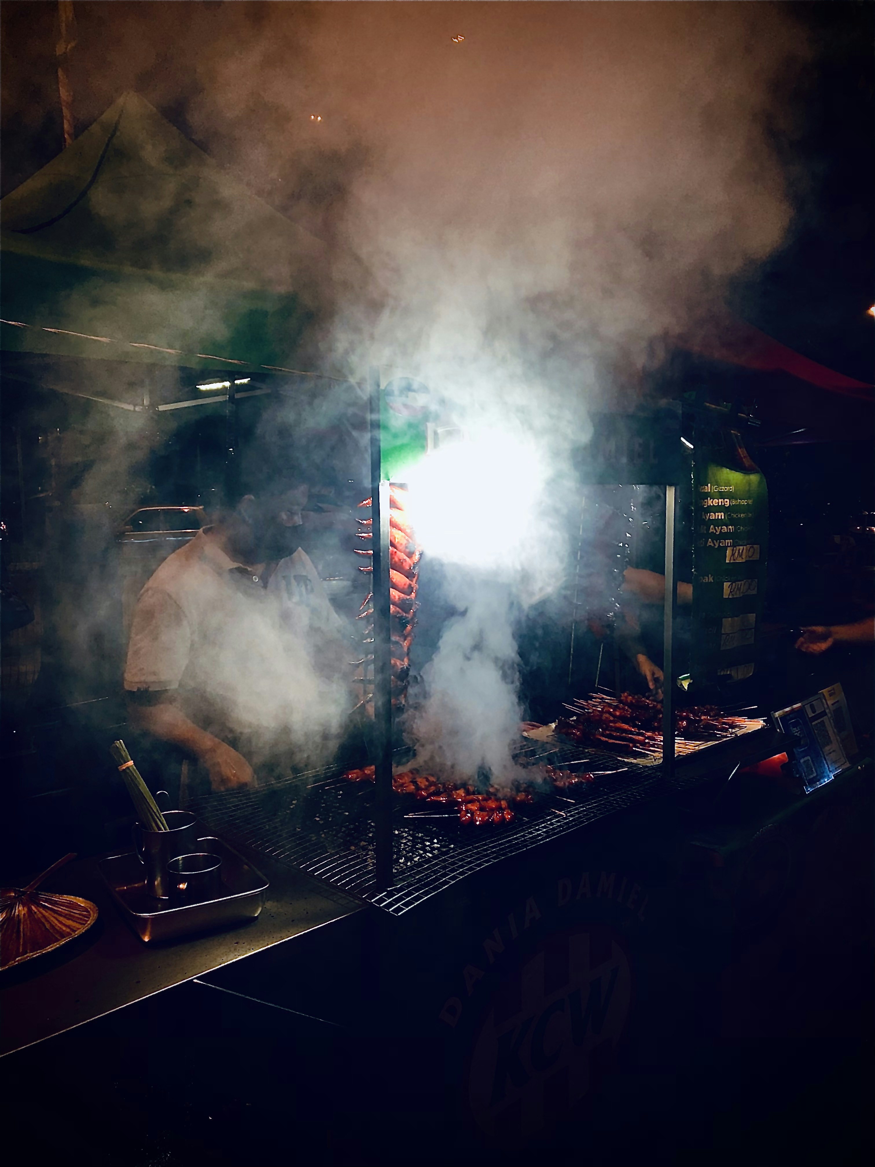 Vibrant street food vendor grilling skewers, enveloped in a cloud of smoke under colorful canopies.