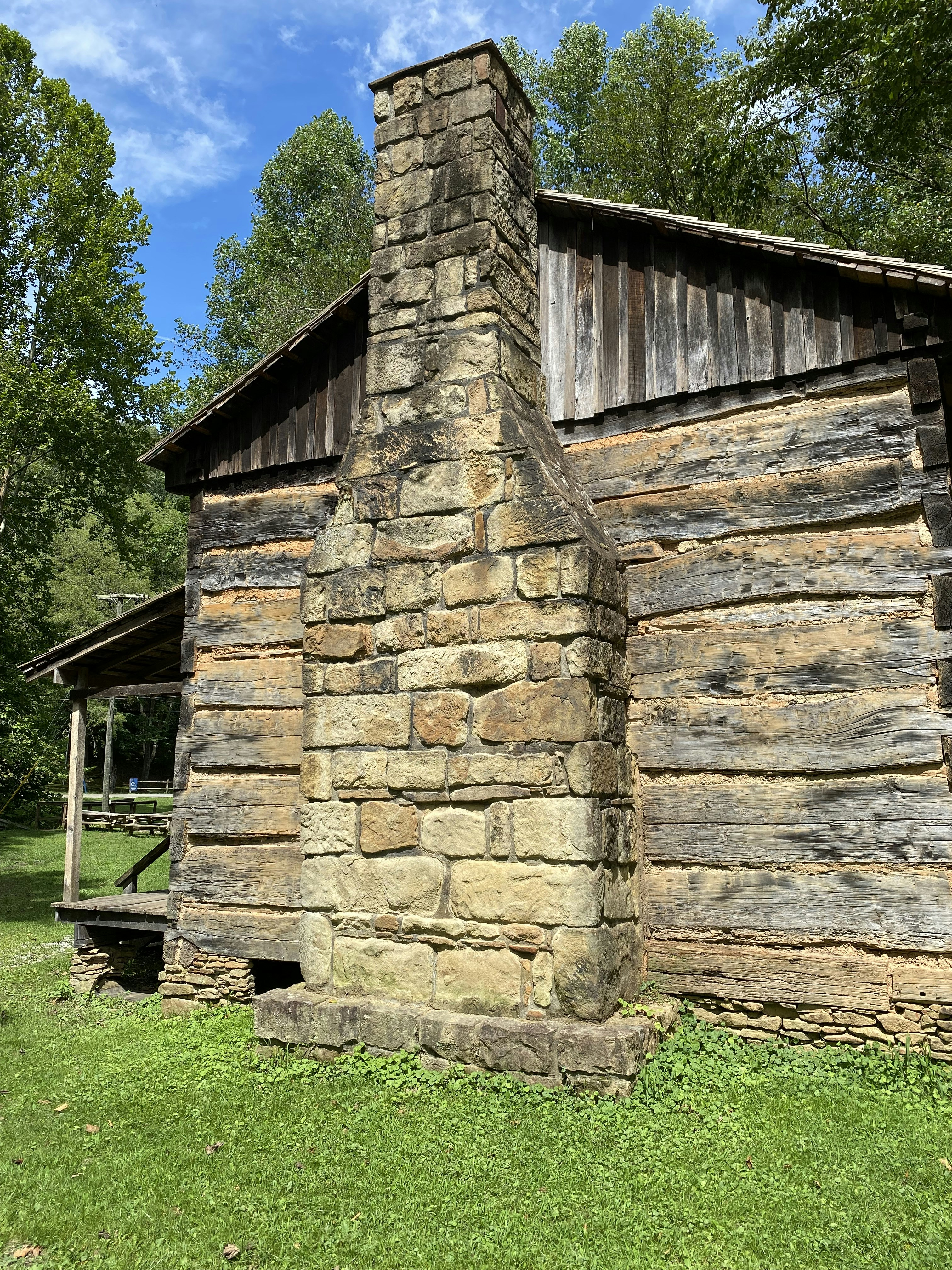 Weathered wooden cabin with a prominent stone chimney surrounded by lush greenery, showcasing traditional craftsmanship and rustic charm.