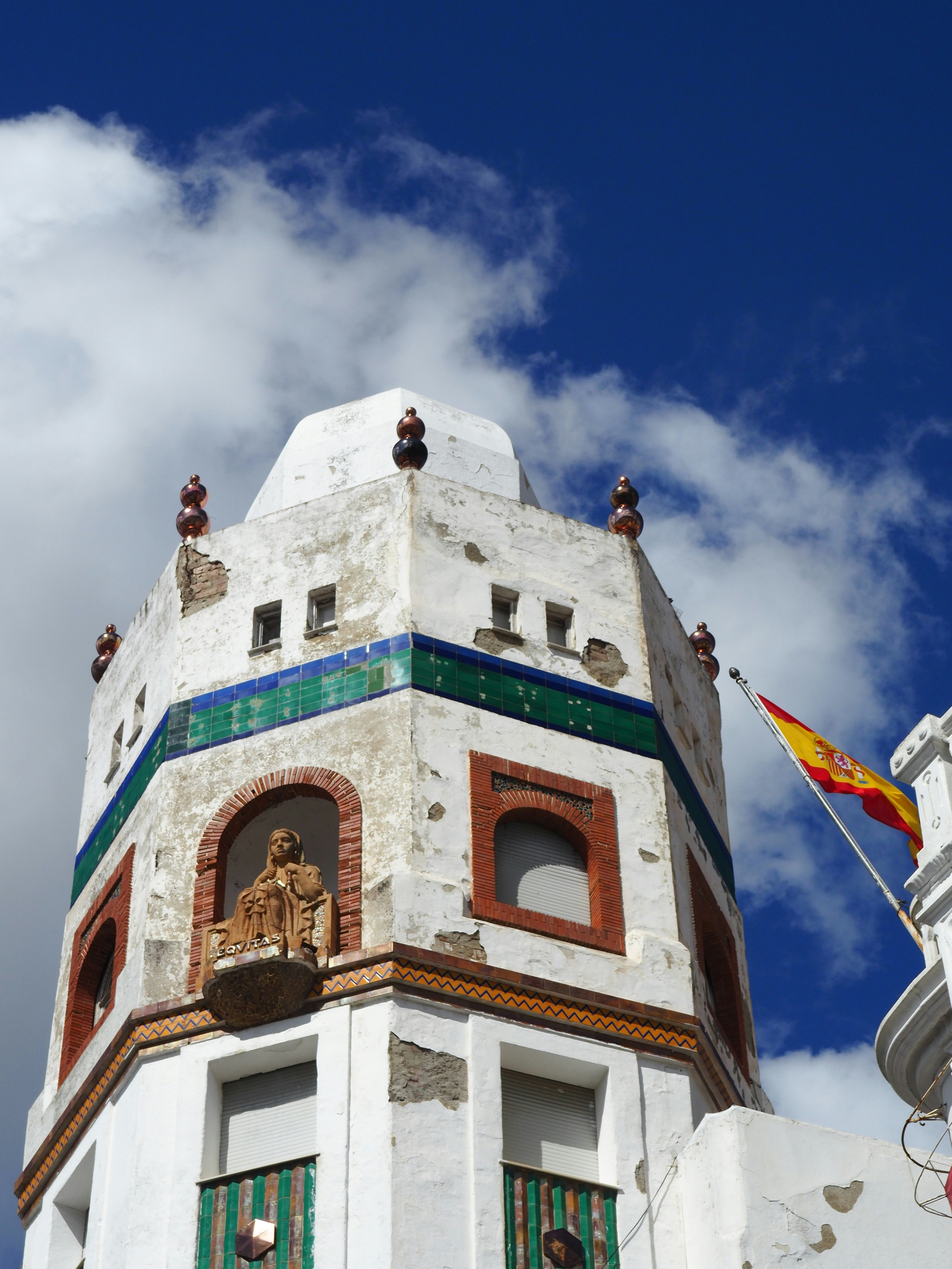 Historic tower adorned with intricate details and a statue, set against a backdrop of dramatic clouds and blue sky.