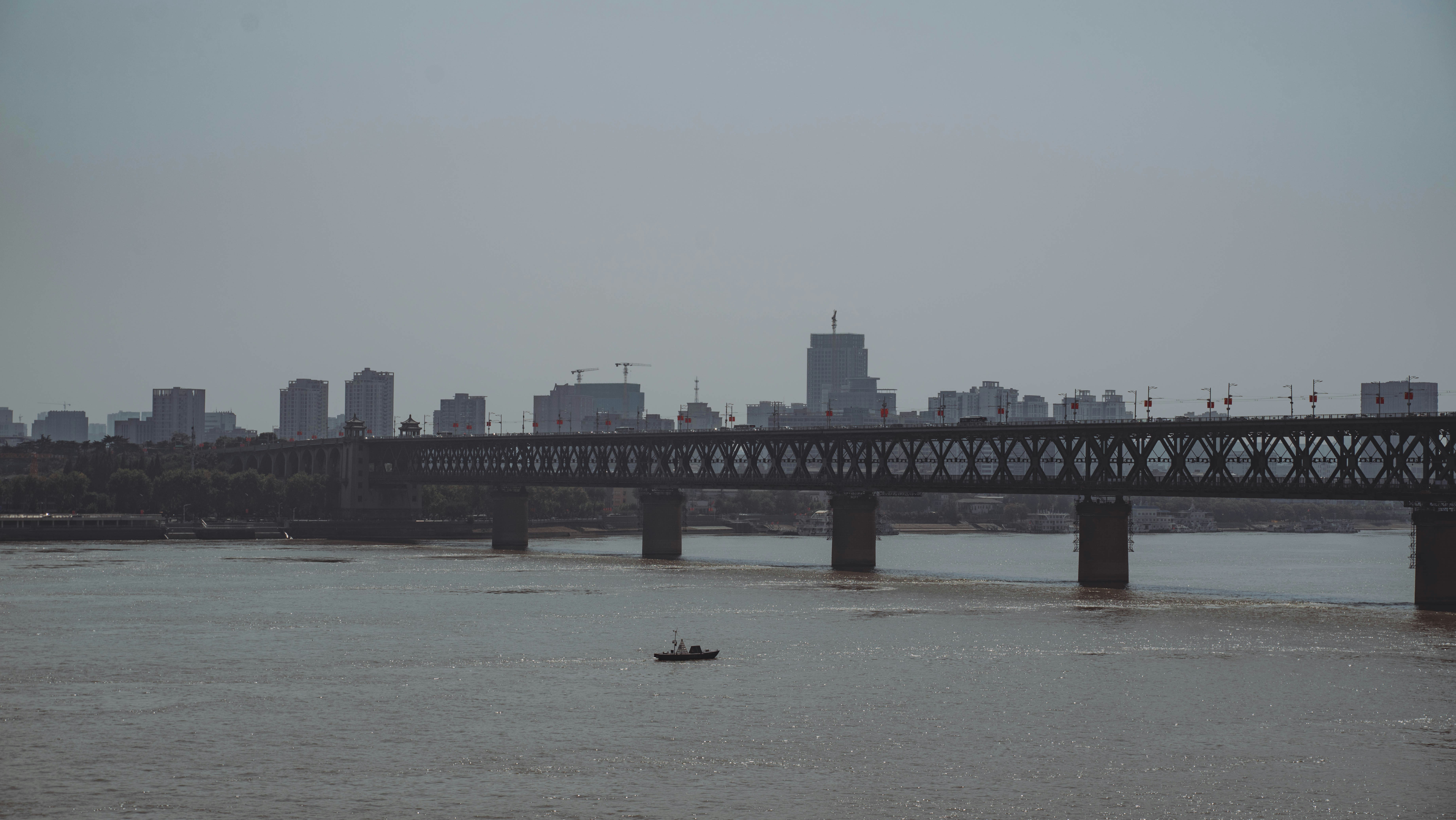 A bridge spans a calm river, with a small boat navigating the water beneath a hazy skyline. The scene captures the harmony between urban architecture and natural elements.