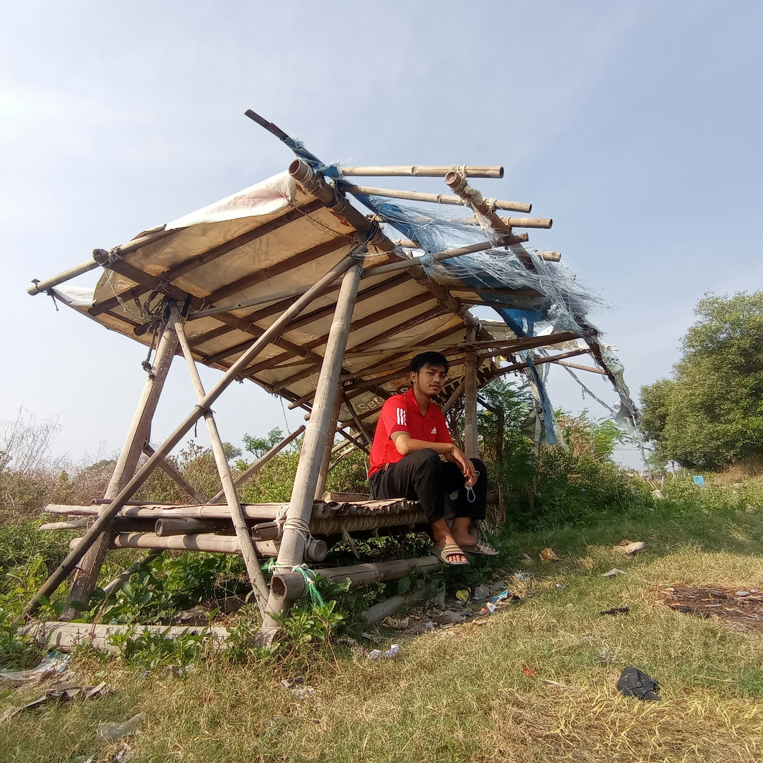 Man sits on a bamboo platform beneath a makeshift shelter built from poles and tarps in a sunlit rural field. The rustic structure and open landscape convey a moment of quiet resilience.