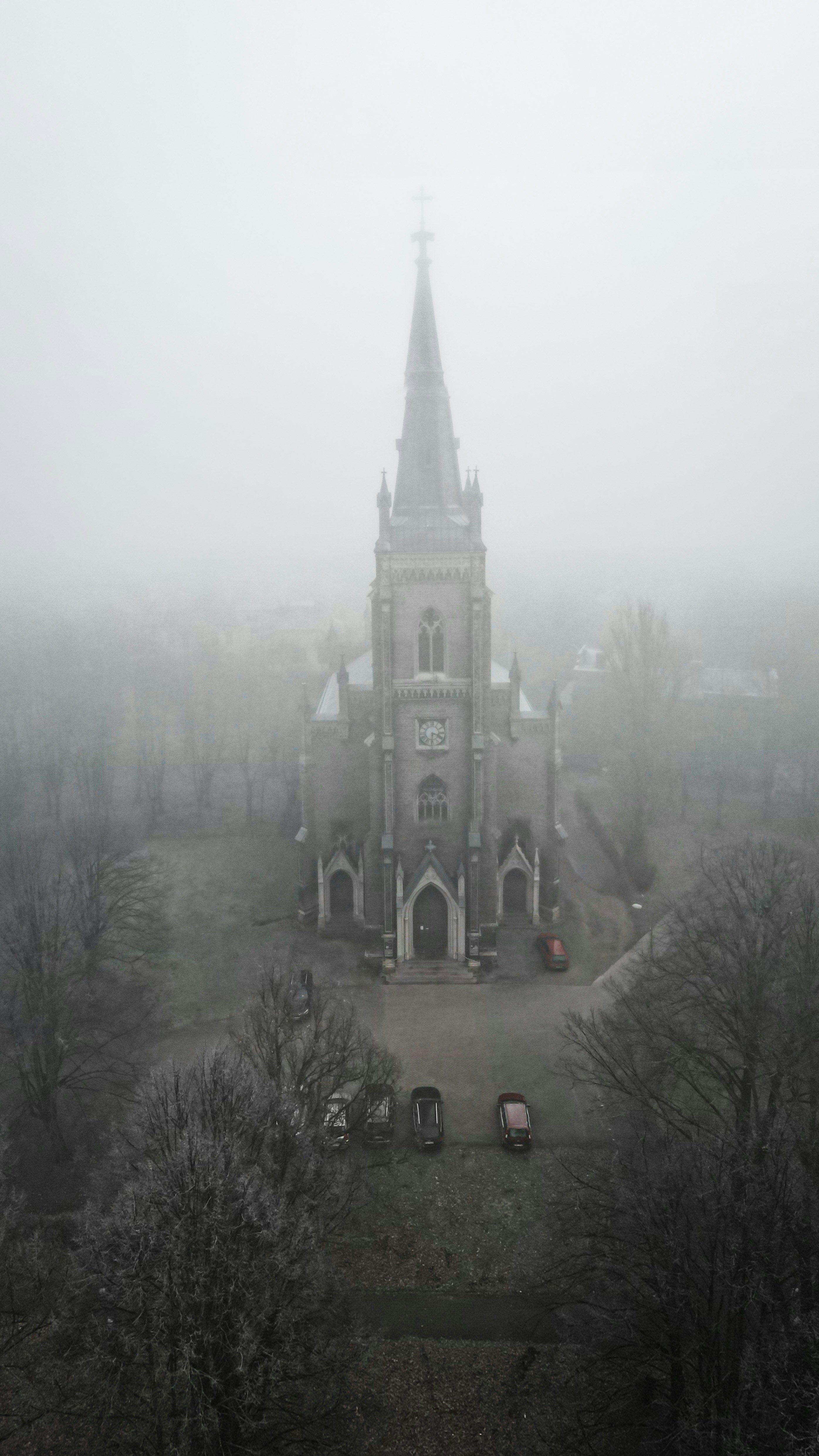 Gothic church emerges through dense fog, surrounded by barren trees and parked cars. The scene exudes an eerie yet serene atmosphere.
