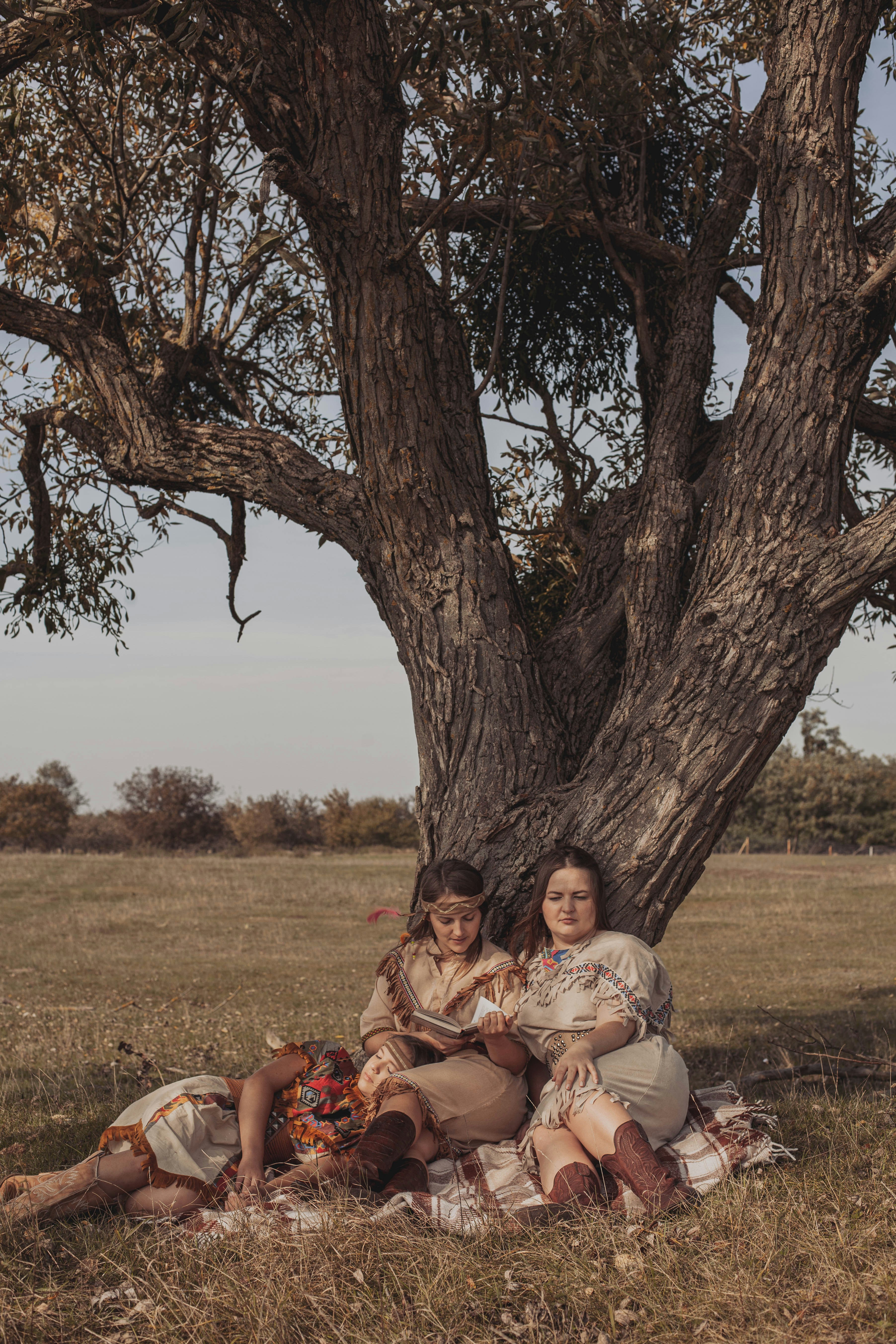 Two Women in Traditional Dress Read Beneath a Sprawling Oak in a Sunlit Prairie
