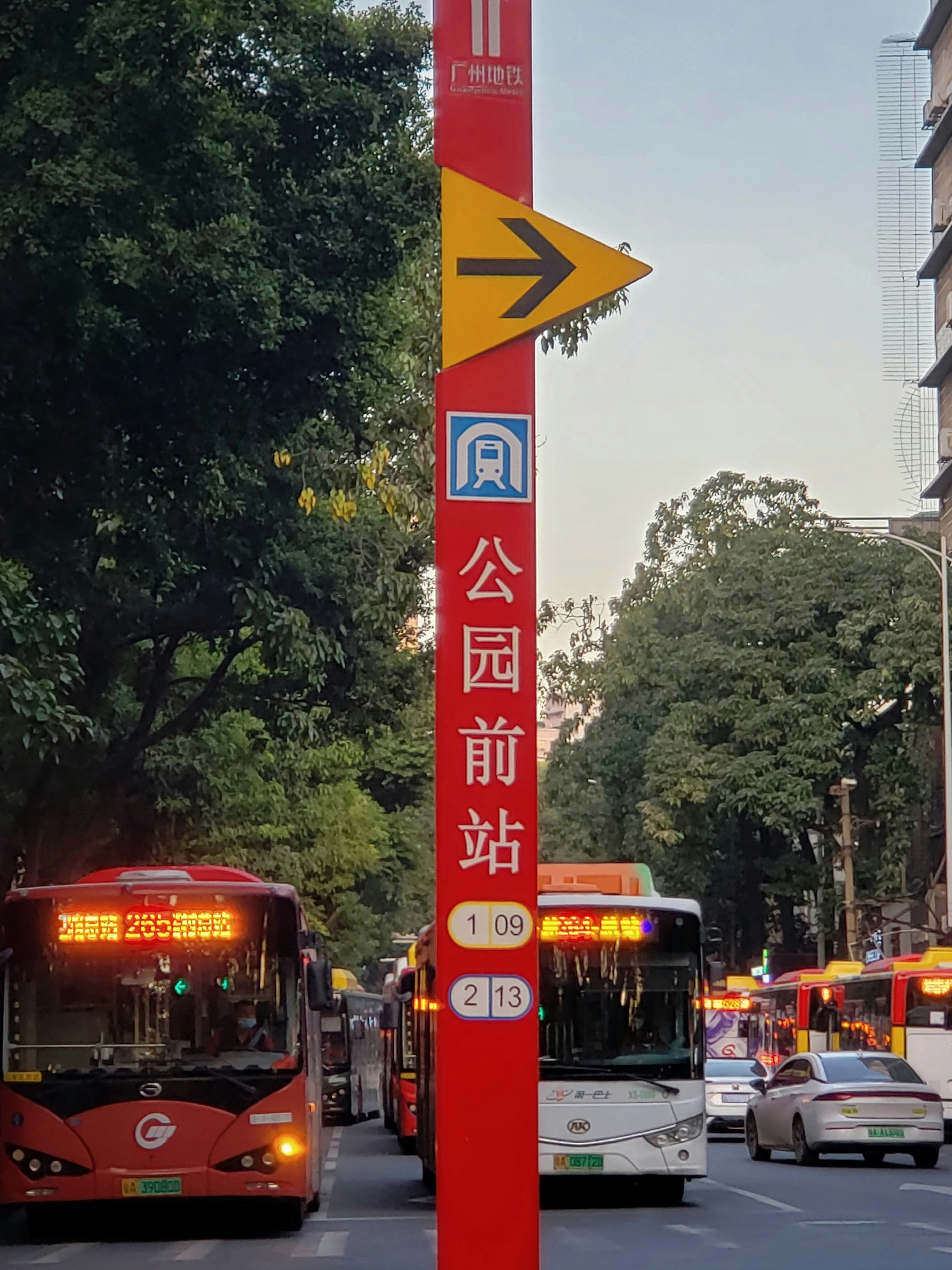 A bus stop sign in a bustling city, featuring route numbers and directional arrows amidst a backdrop of urban greenery and public transport vehicles.