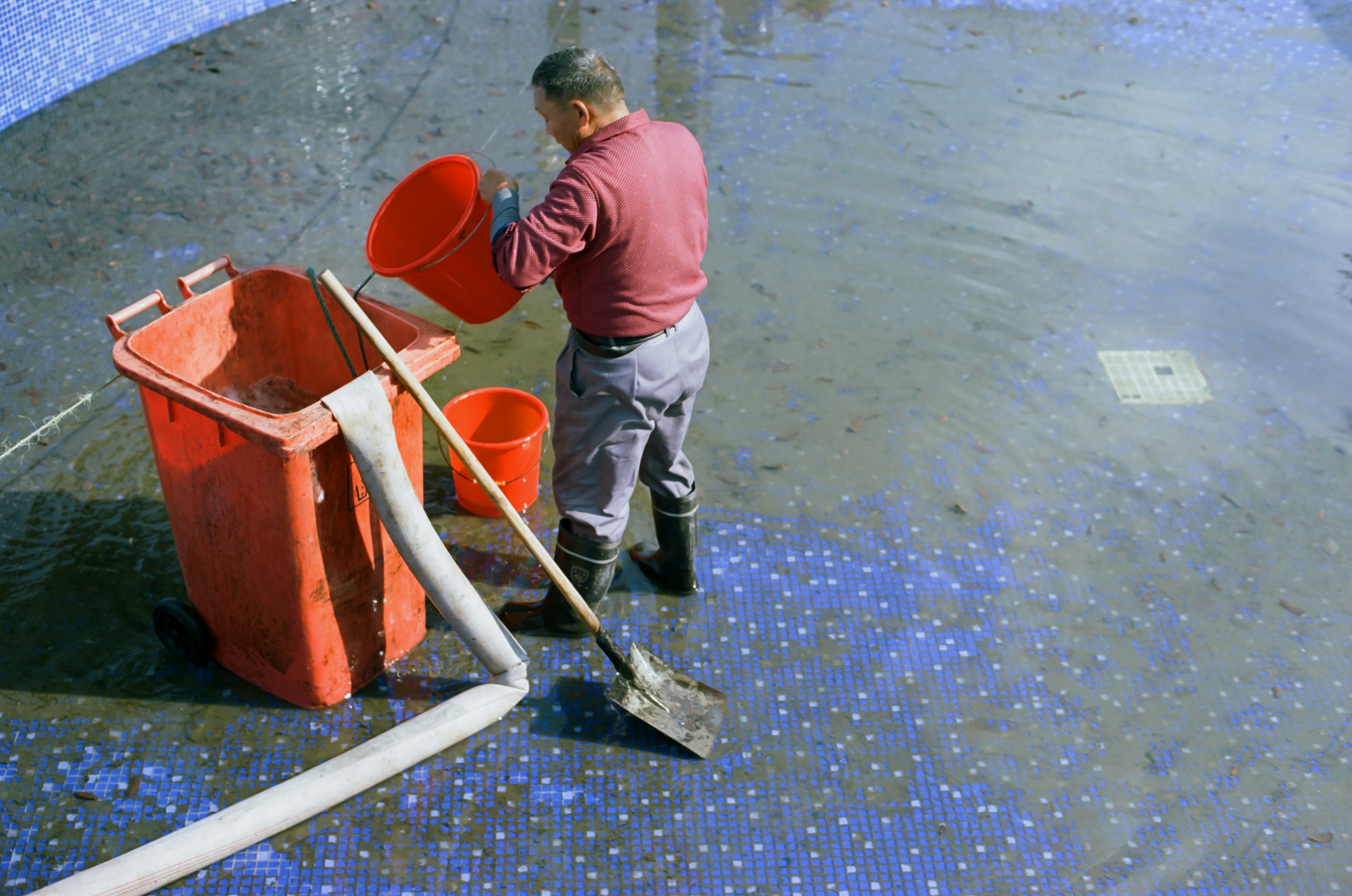 A worker in a magenta shirt handles a red bucket beside a large red bin on a wet blue-tiled surface, with a hose and a floor drain nearby.