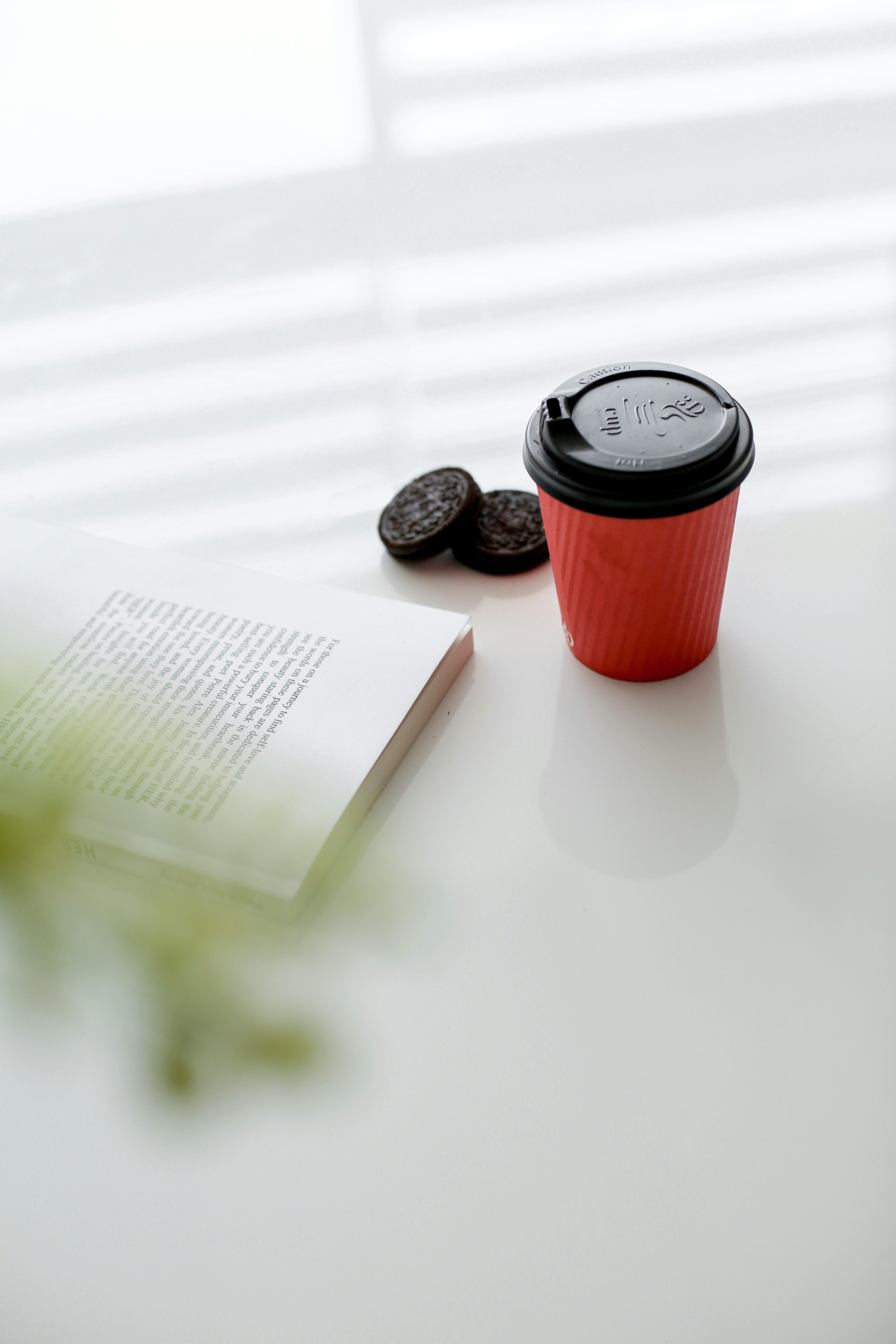 A vibrant red coffee cup sits beside an open book and chocolate cookies on a glossy surface, illuminated by soft natural light.