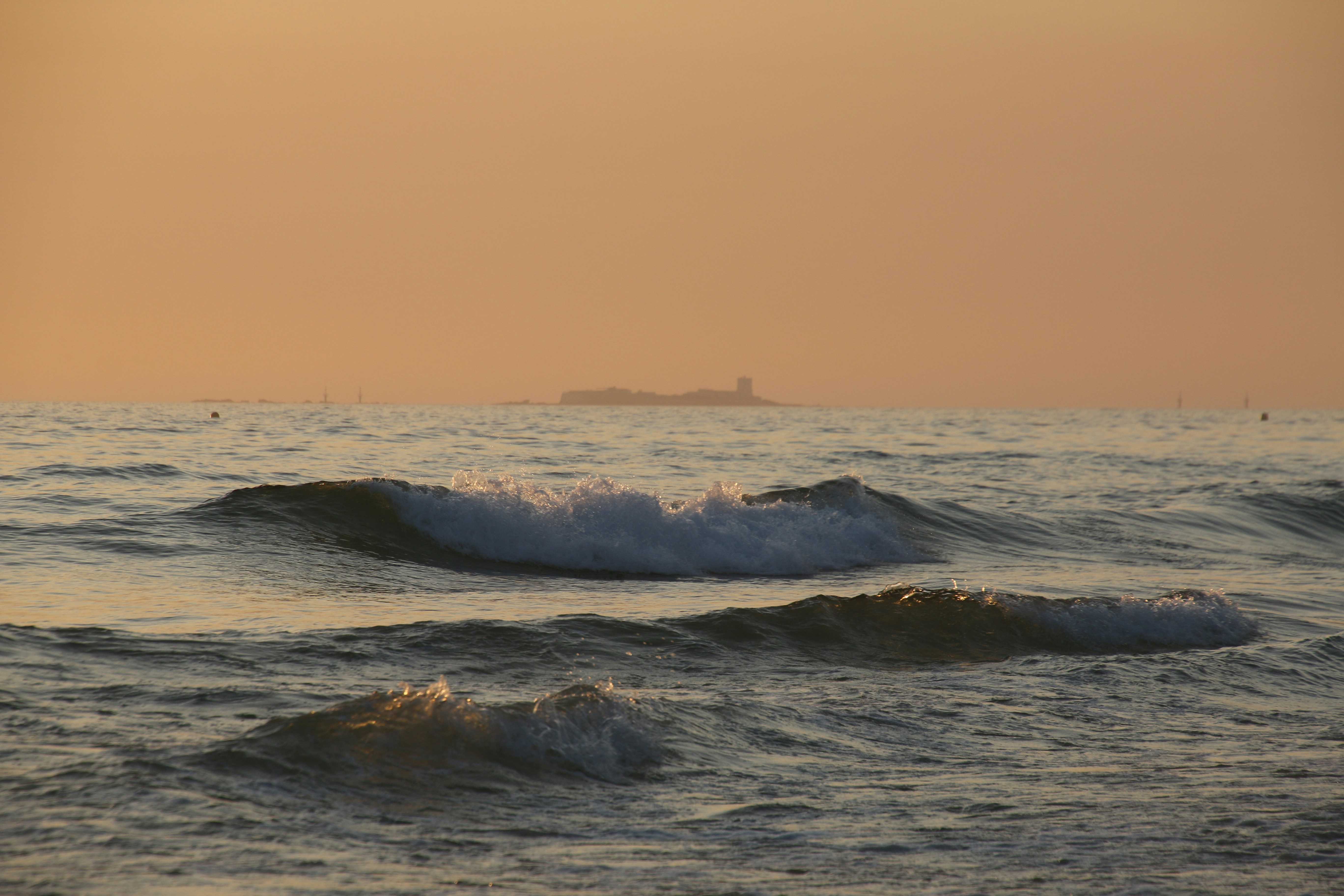 Gentle waves roll under a warm, orange sky with a distant silhouette on the horizon.