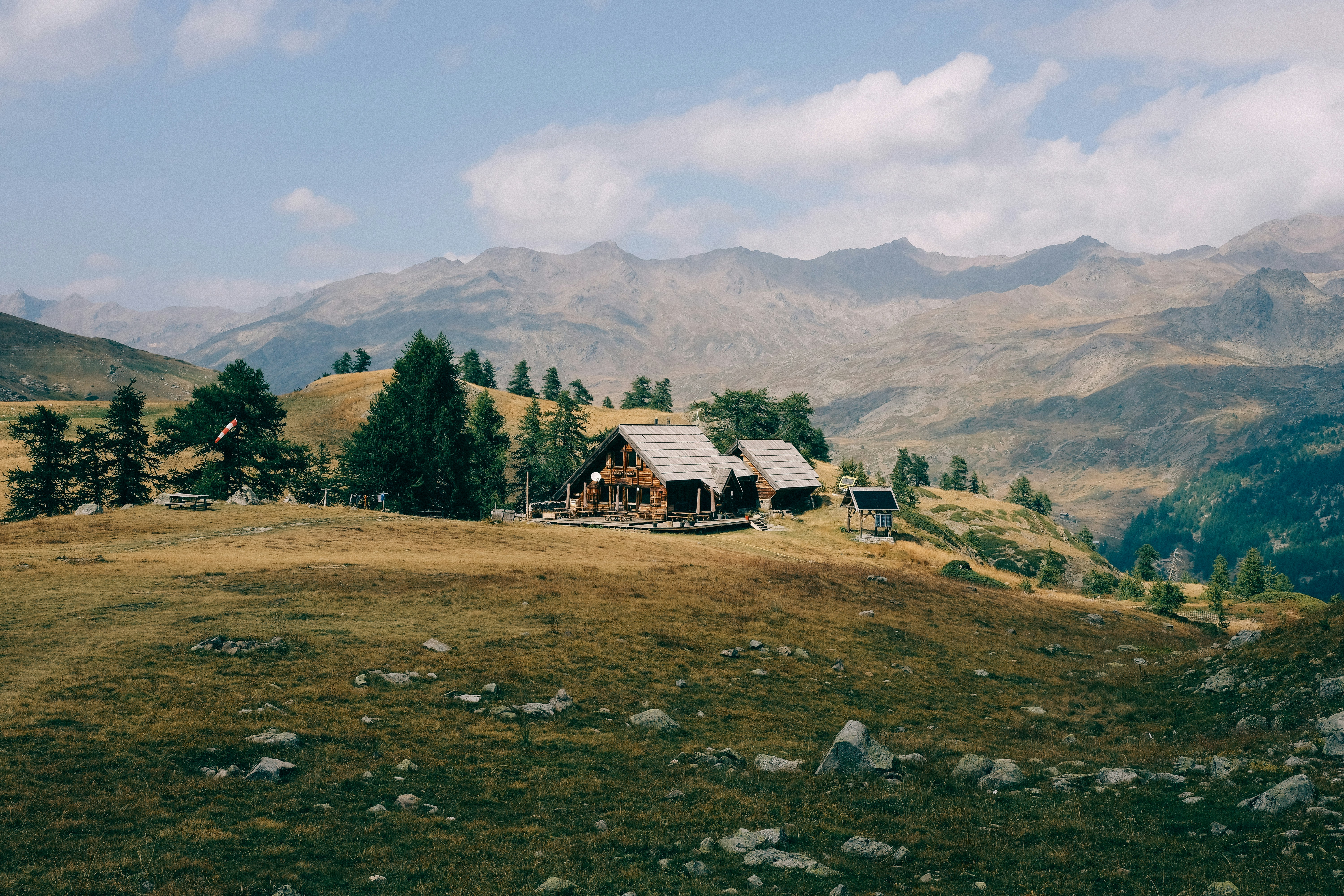 Wooden cabin nestled in a serene alpine landscape, surrounded by rolling hills and towering mountains under a partly cloudy sky.