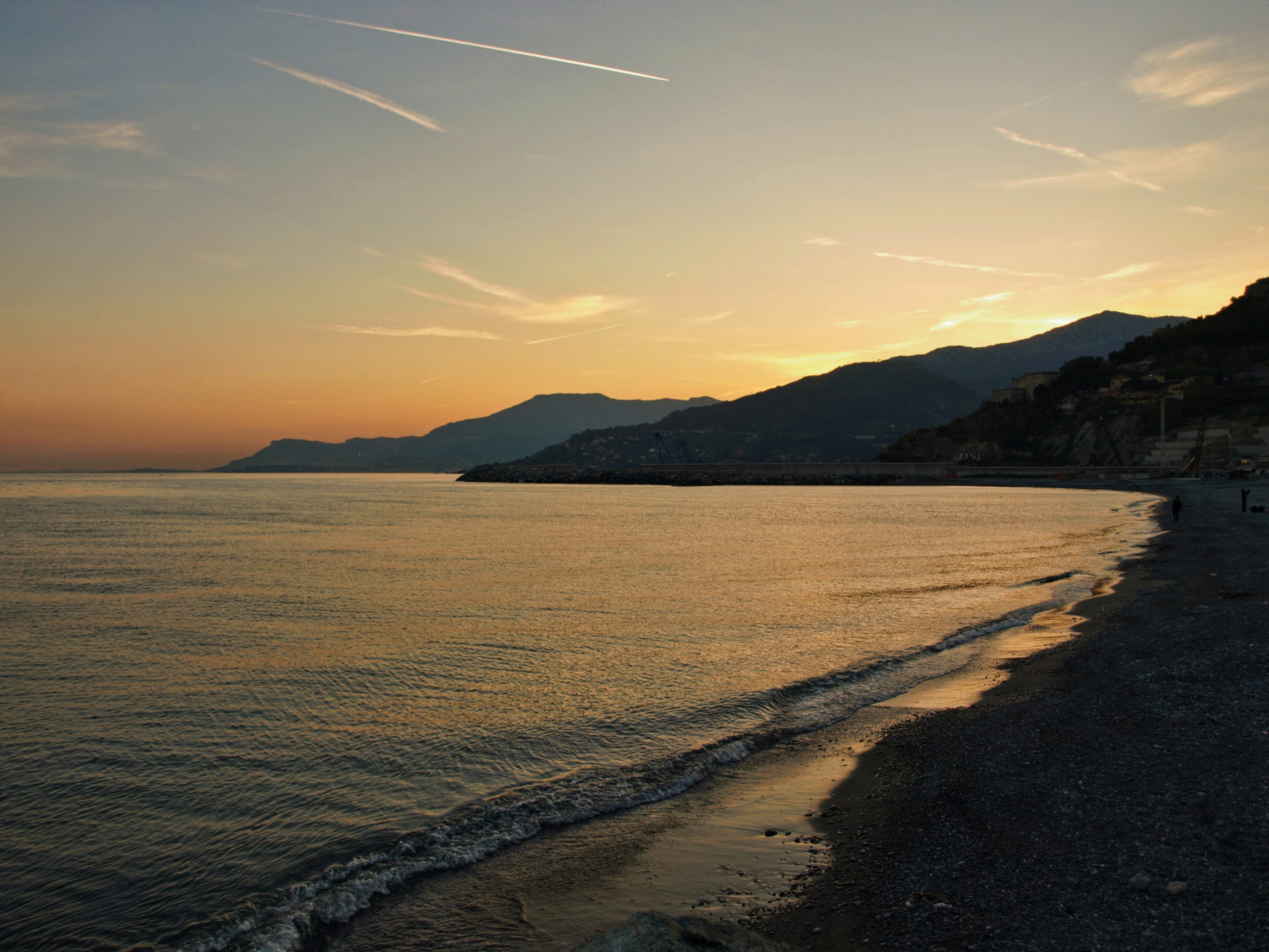 Sunset casting golden hues over calm ocean waves with distant mountains silhouetted against the sky.