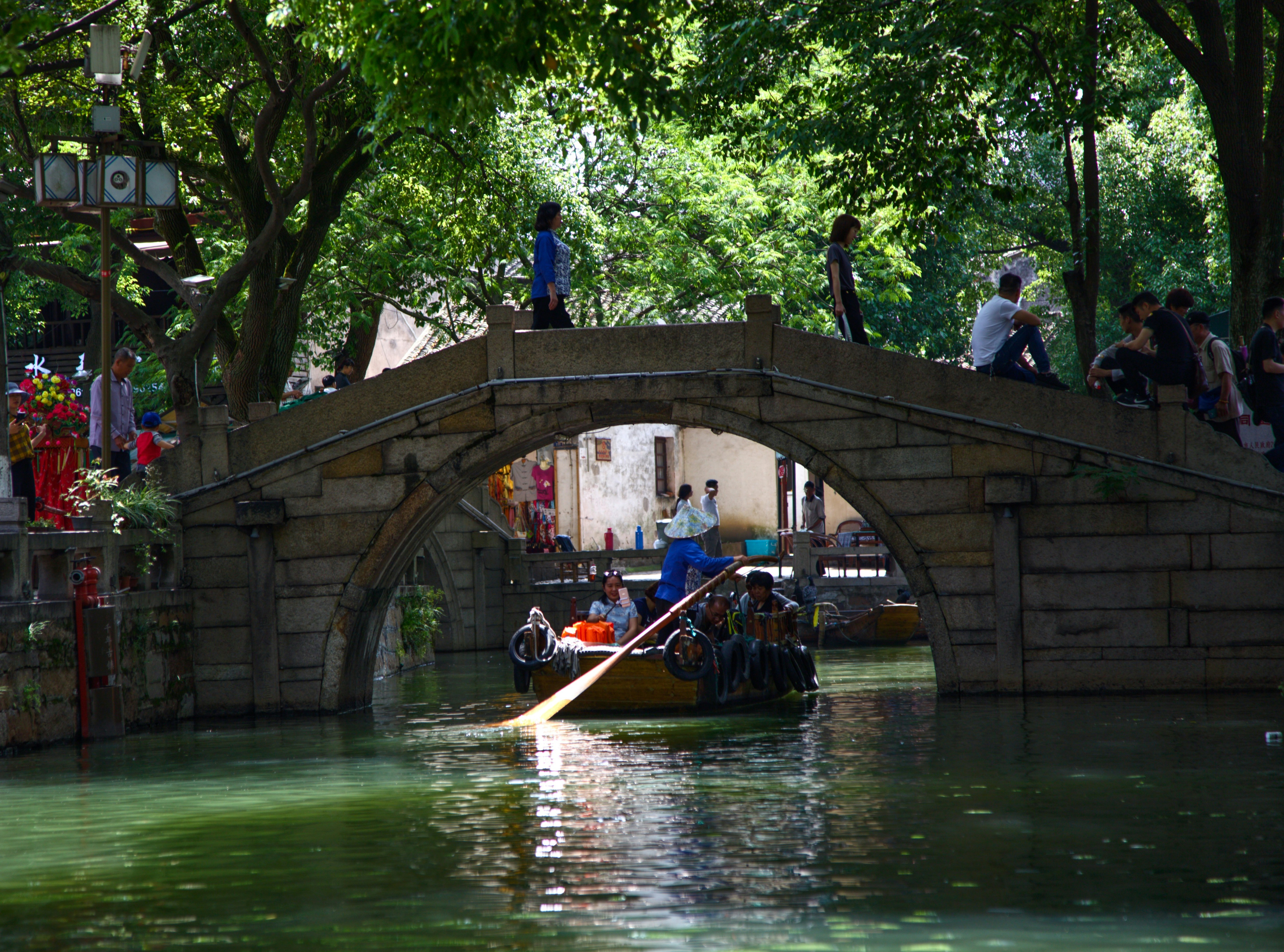 Wooden boat gliding under a stone bridge in a lush, tree-lined canal of Tongli water town.