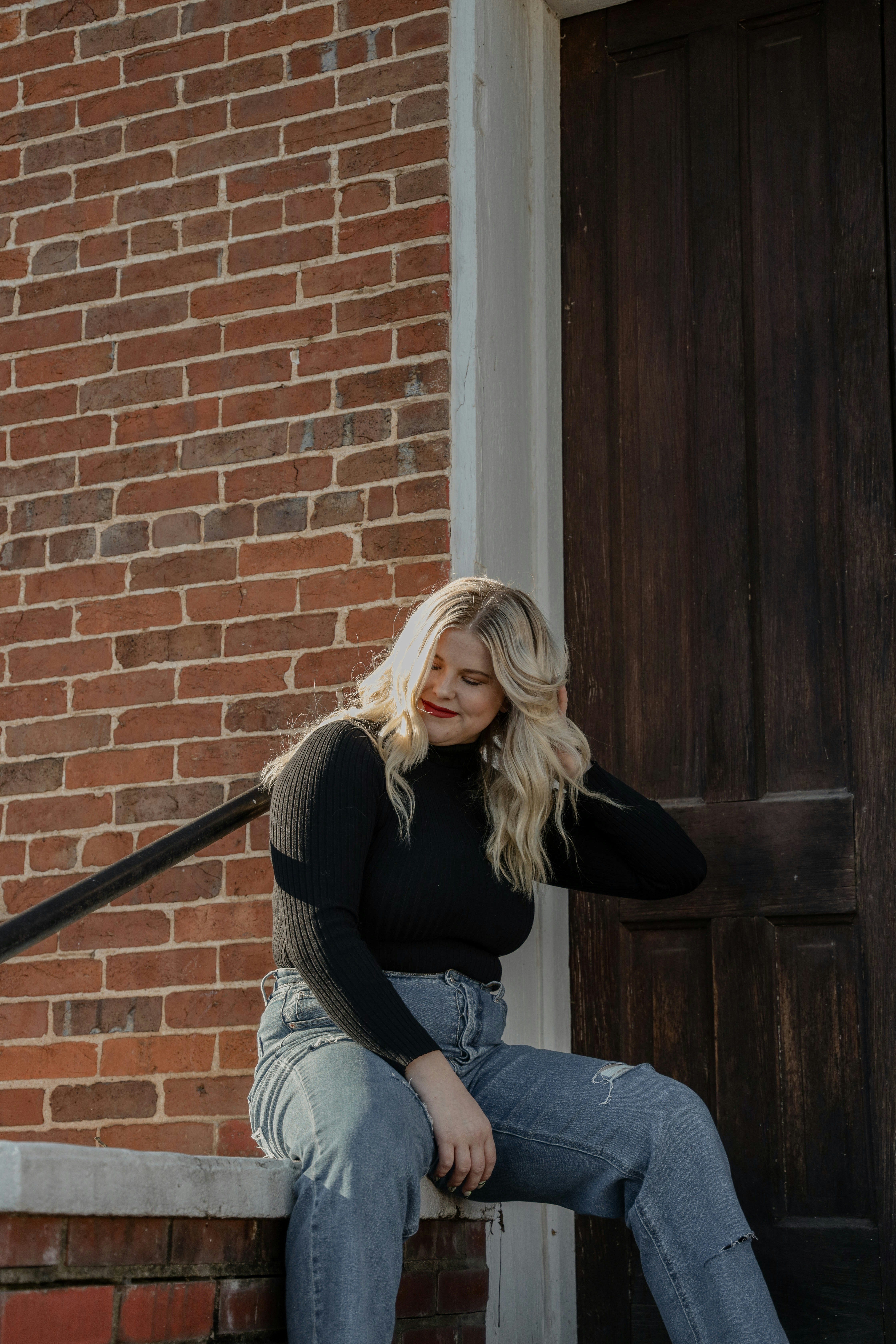 A young woman with wavy blonde hair sits casually on a brick step, adorned in a black sweater and light jeans, against a rustic wooden door.