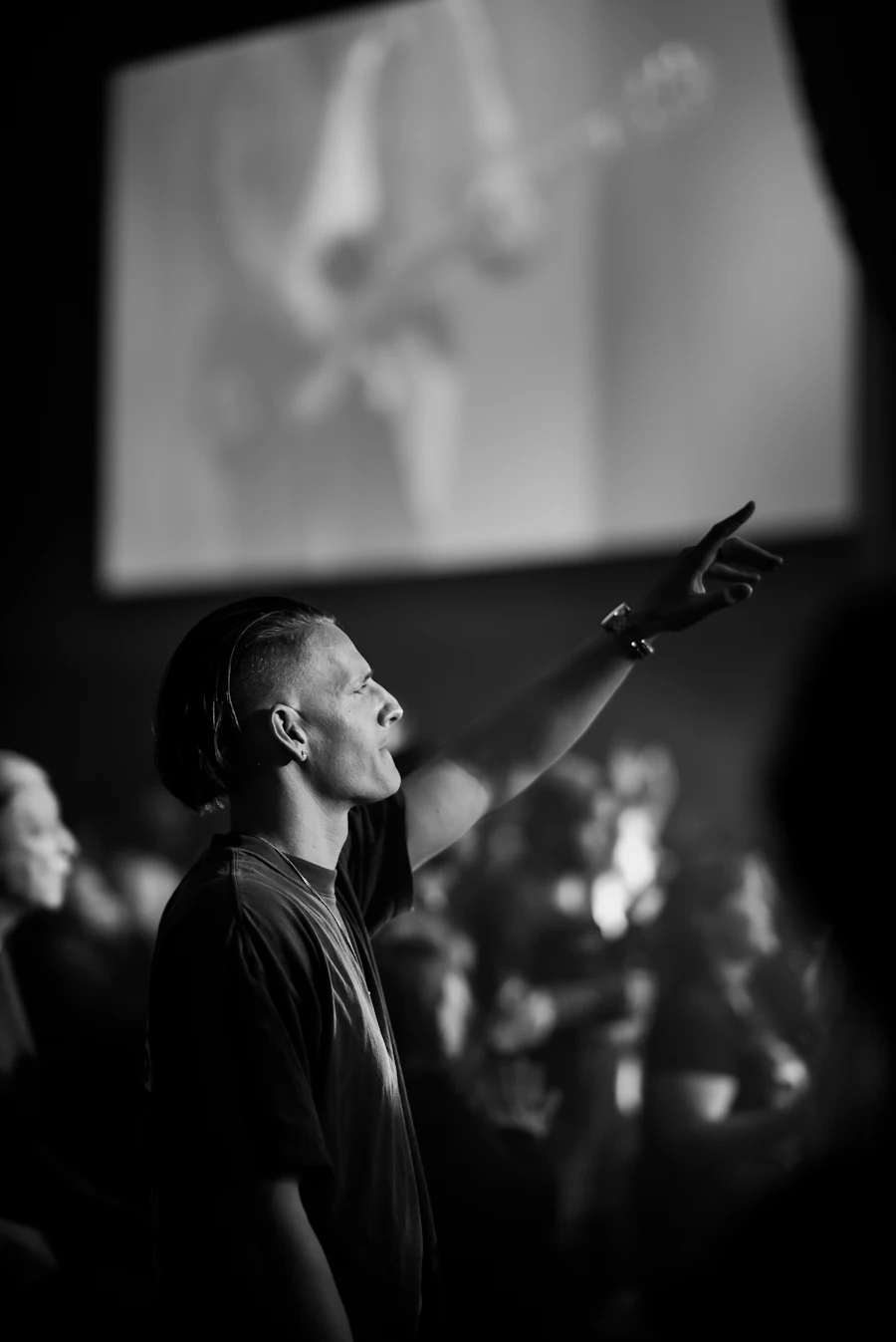 Black-and-white scene of a man in profile raising his arm toward a stage or screen, with blurred crowd and projection in the background. Moody low-light atmosphere with strong directional contrast.