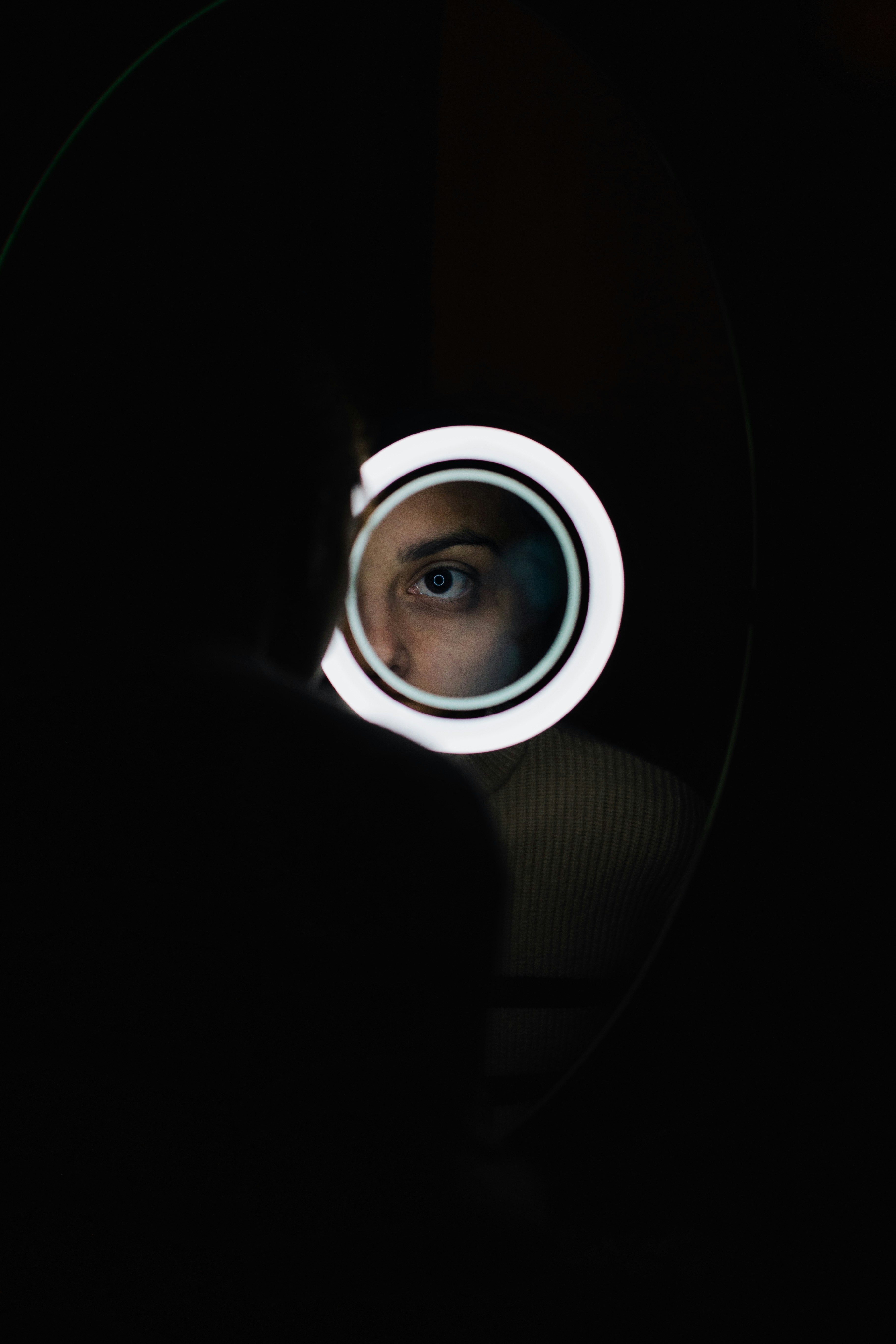 A person gazes into a circular mirror with a soft glow, revealing a striking blue eye against a dark backdrop.