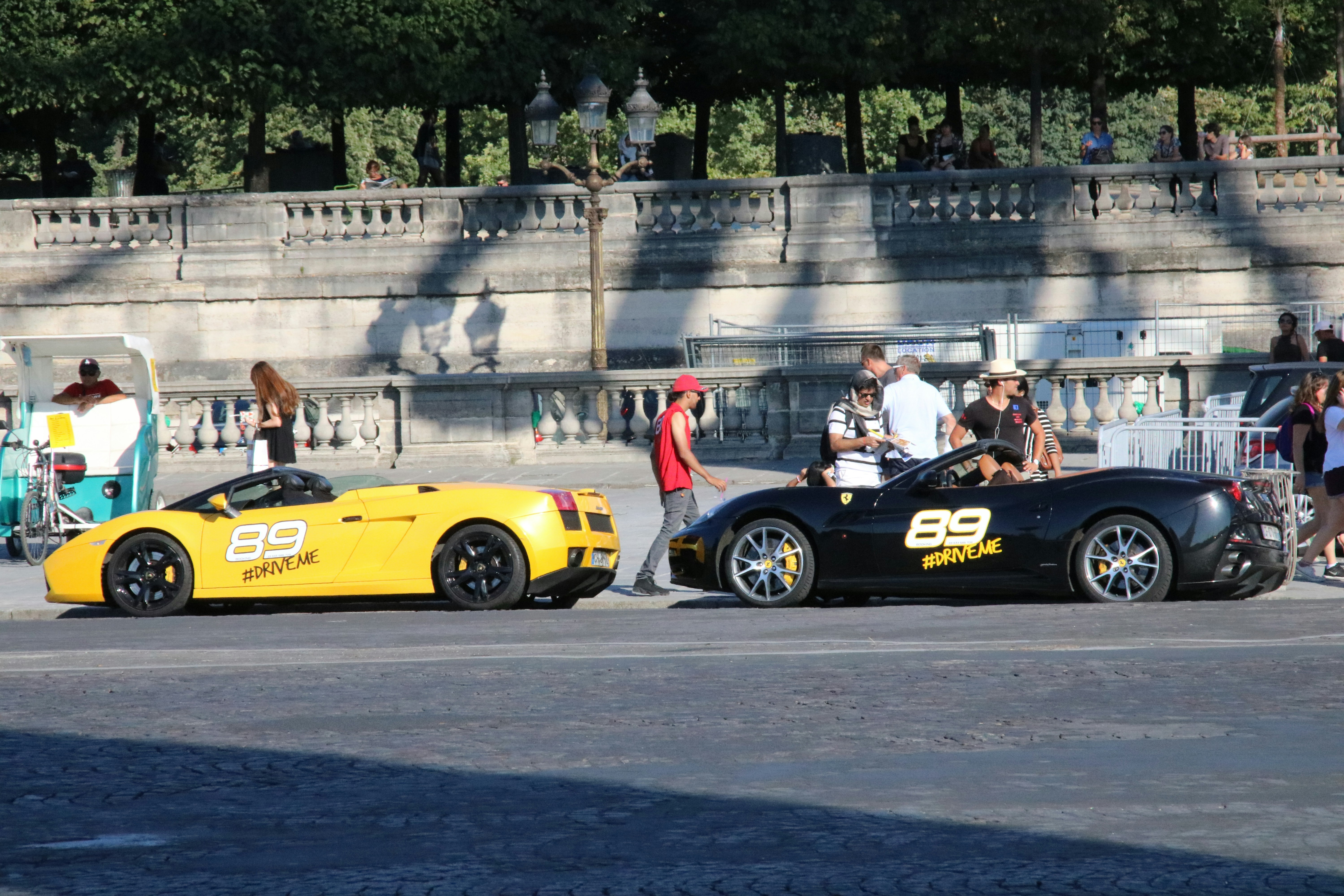 Two luxury sports cars, one yellow and one black, parked side by side in a vibrant urban setting filled with onlookers.