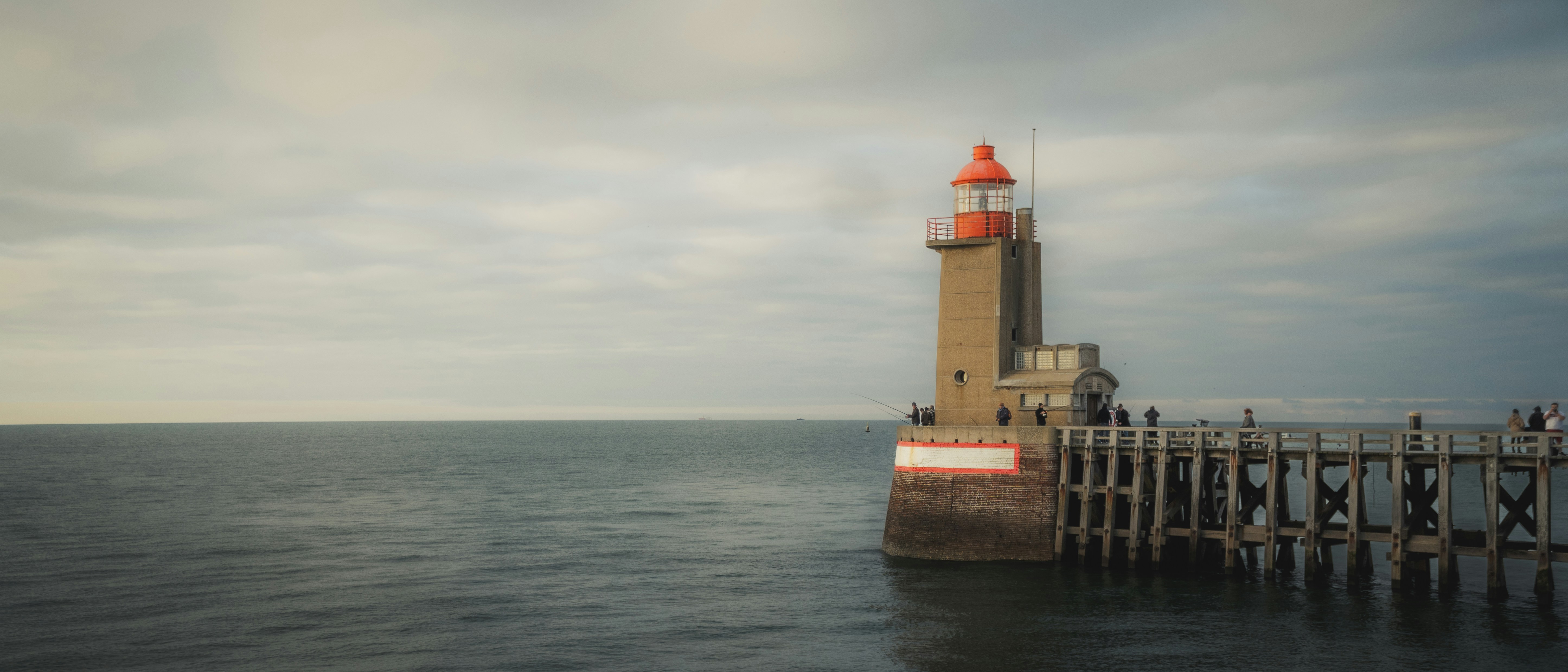 A lighthouse standing tall on a pier, overlooking calm waters under a cloudy sky. The structure features a vibrant orange top, contrasting with the muted tones of the surroundings.