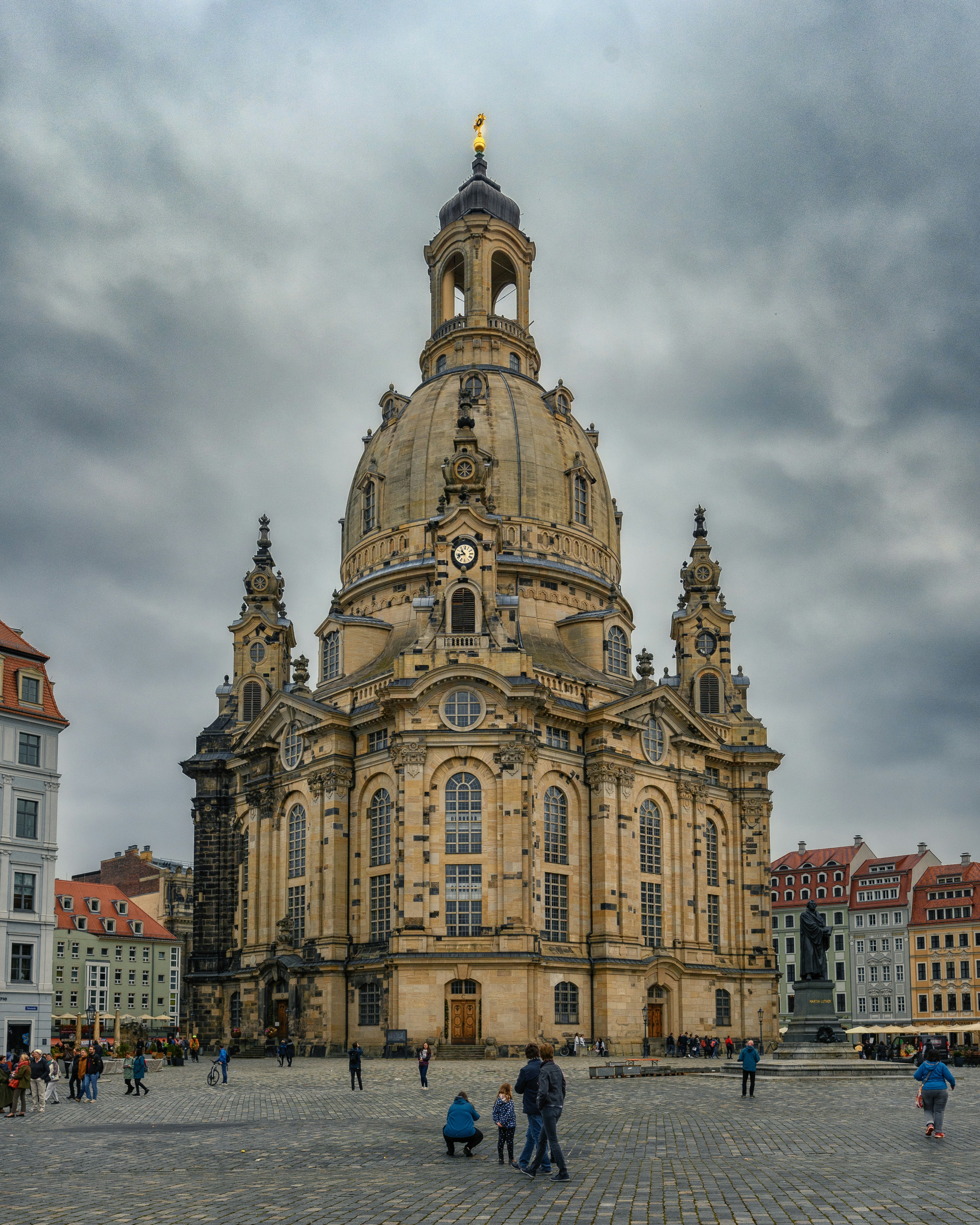Baroque church featuring a prominent dome and intricate stonework, surrounded by visitors in a bustling square.