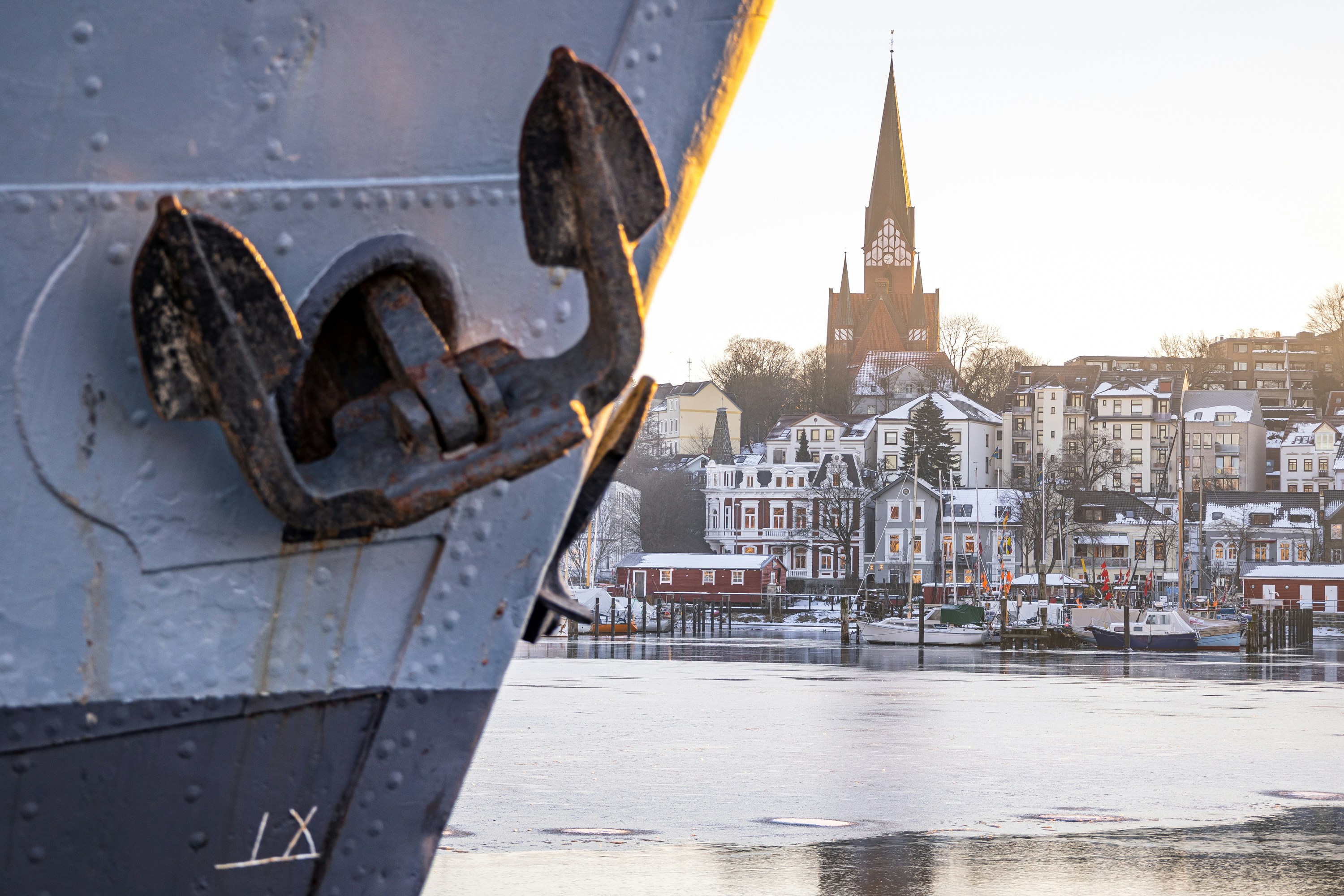 Large ship anchor in foreground with a serene cityscape and church spire across a calm, icy river.