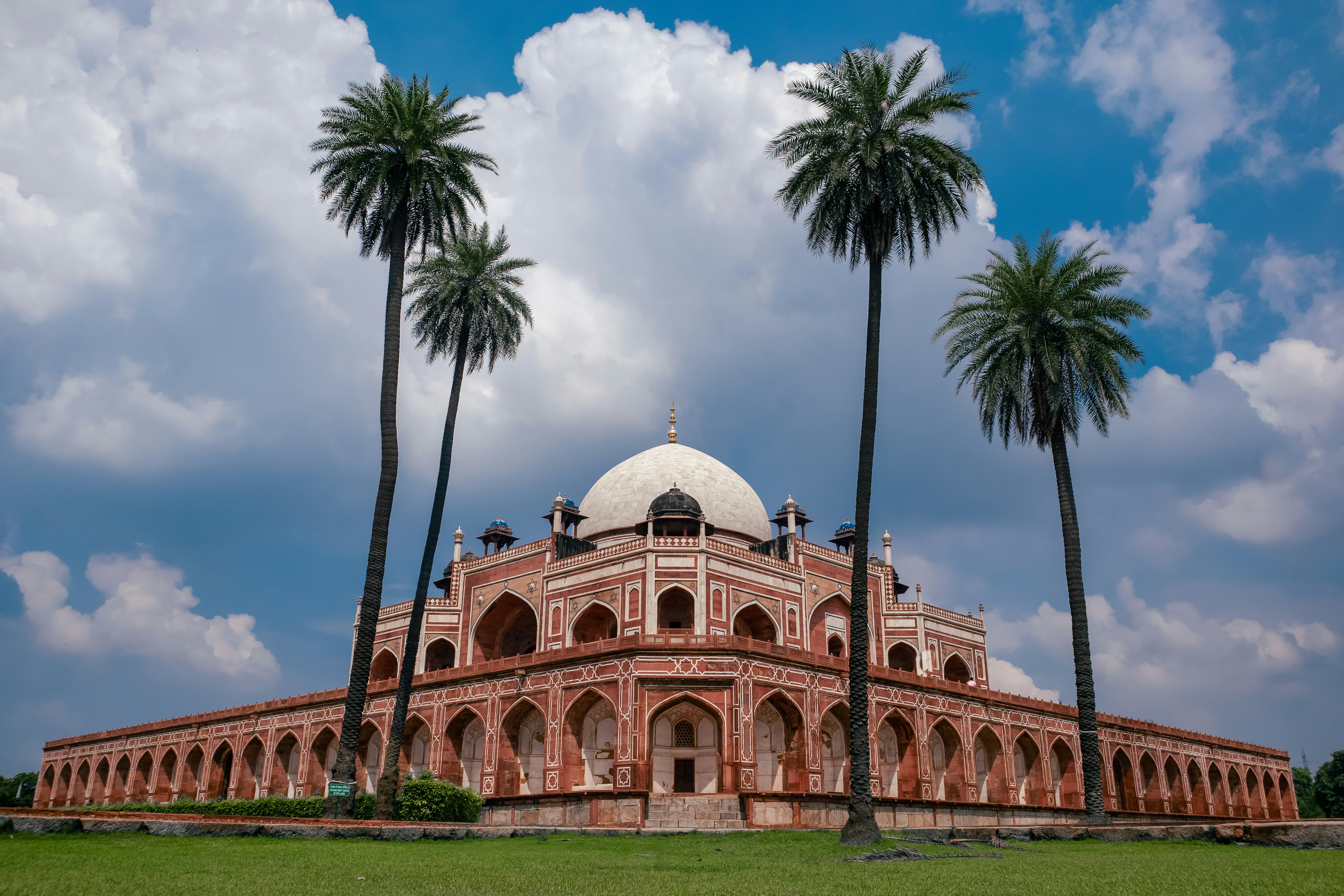 Humayun's Tomb, an iconic example of Mughal architecture, stands surrounded by lush greenery and palm trees under a dramatic sky.