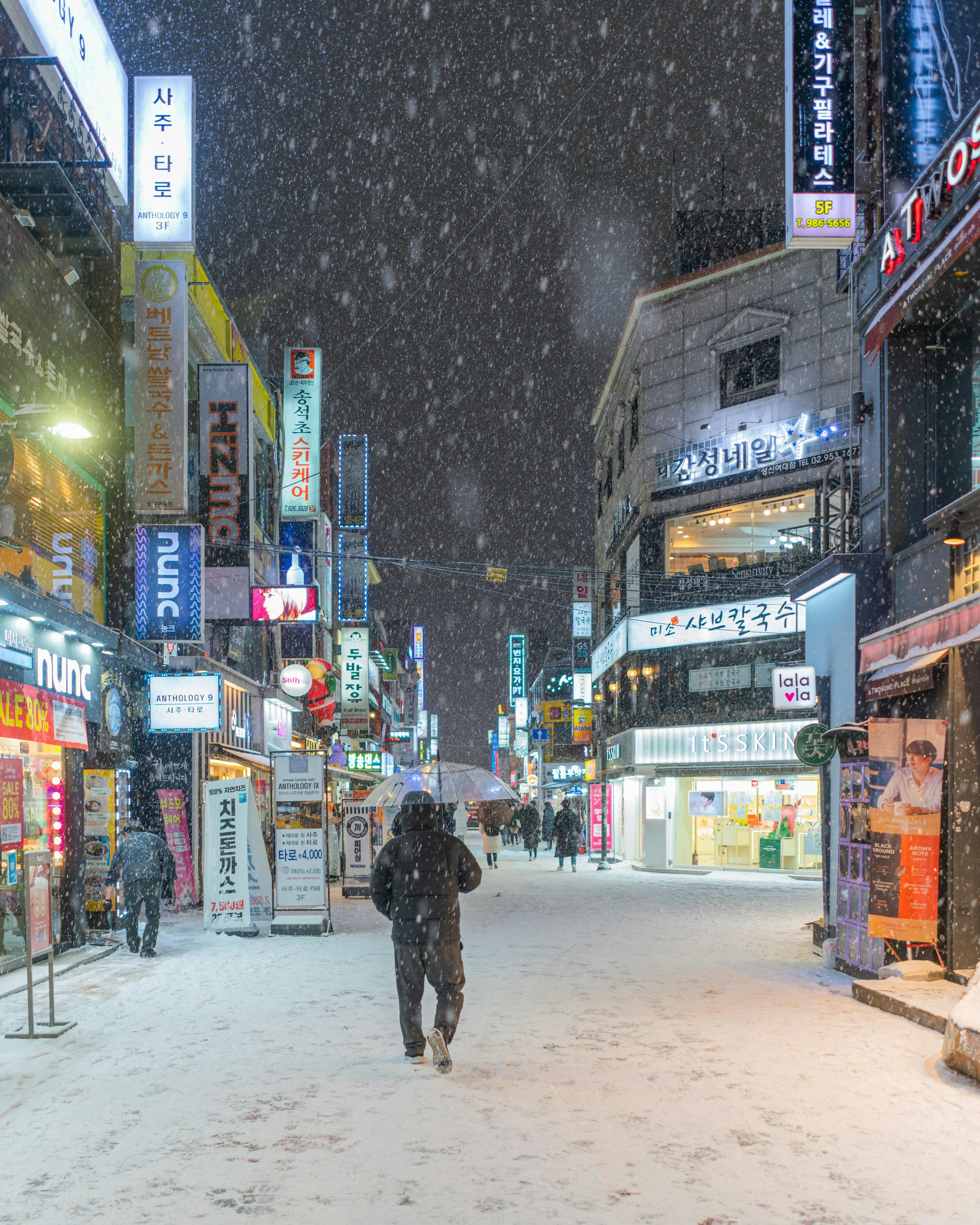 Neon Snowfall on a Neon-Kissed Korean Street: A lone figure in drifting flurries