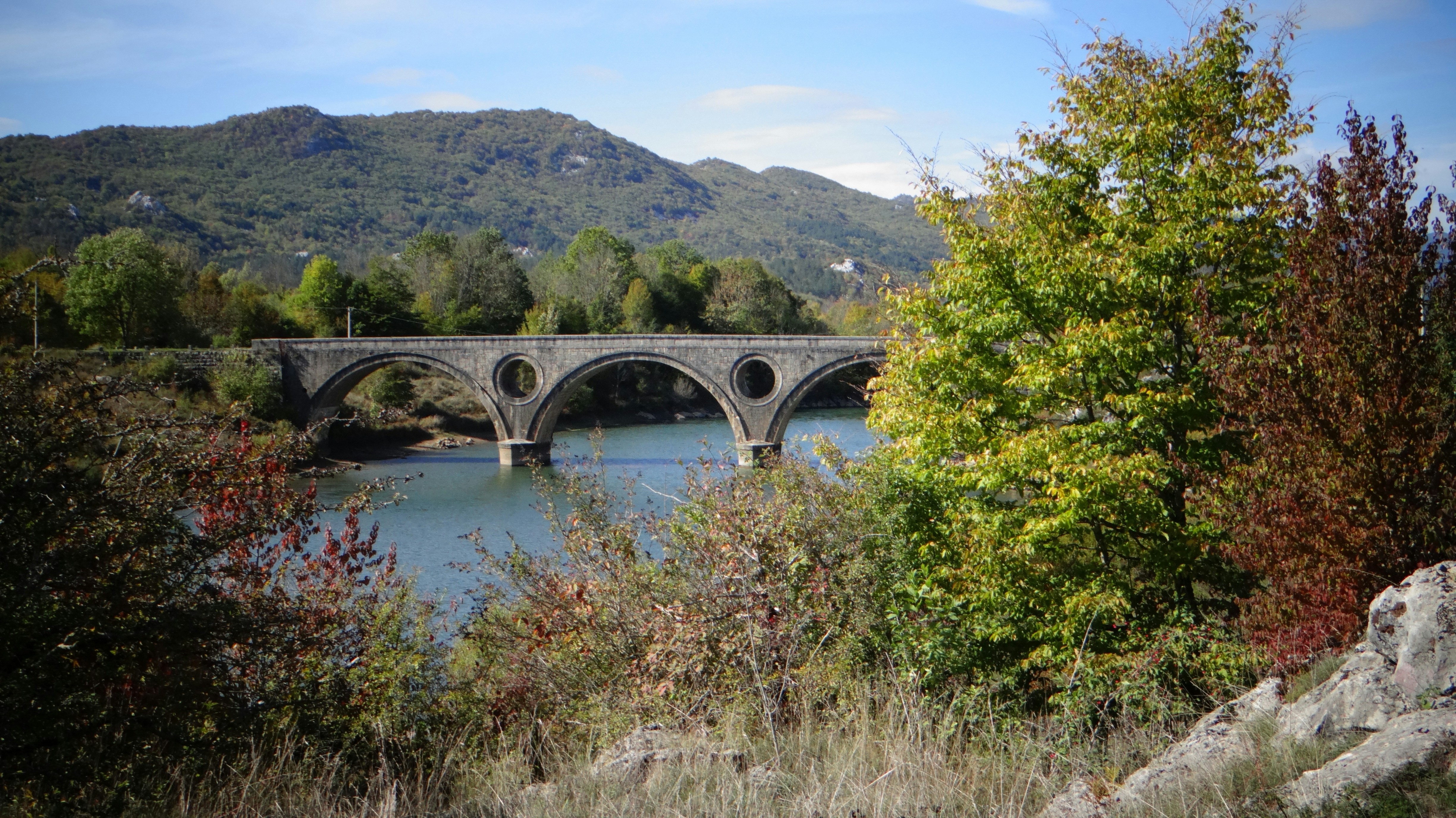 Historic stone bridge spans a serene river, framed by vibrant autumn foliage and rolling hills in the background.