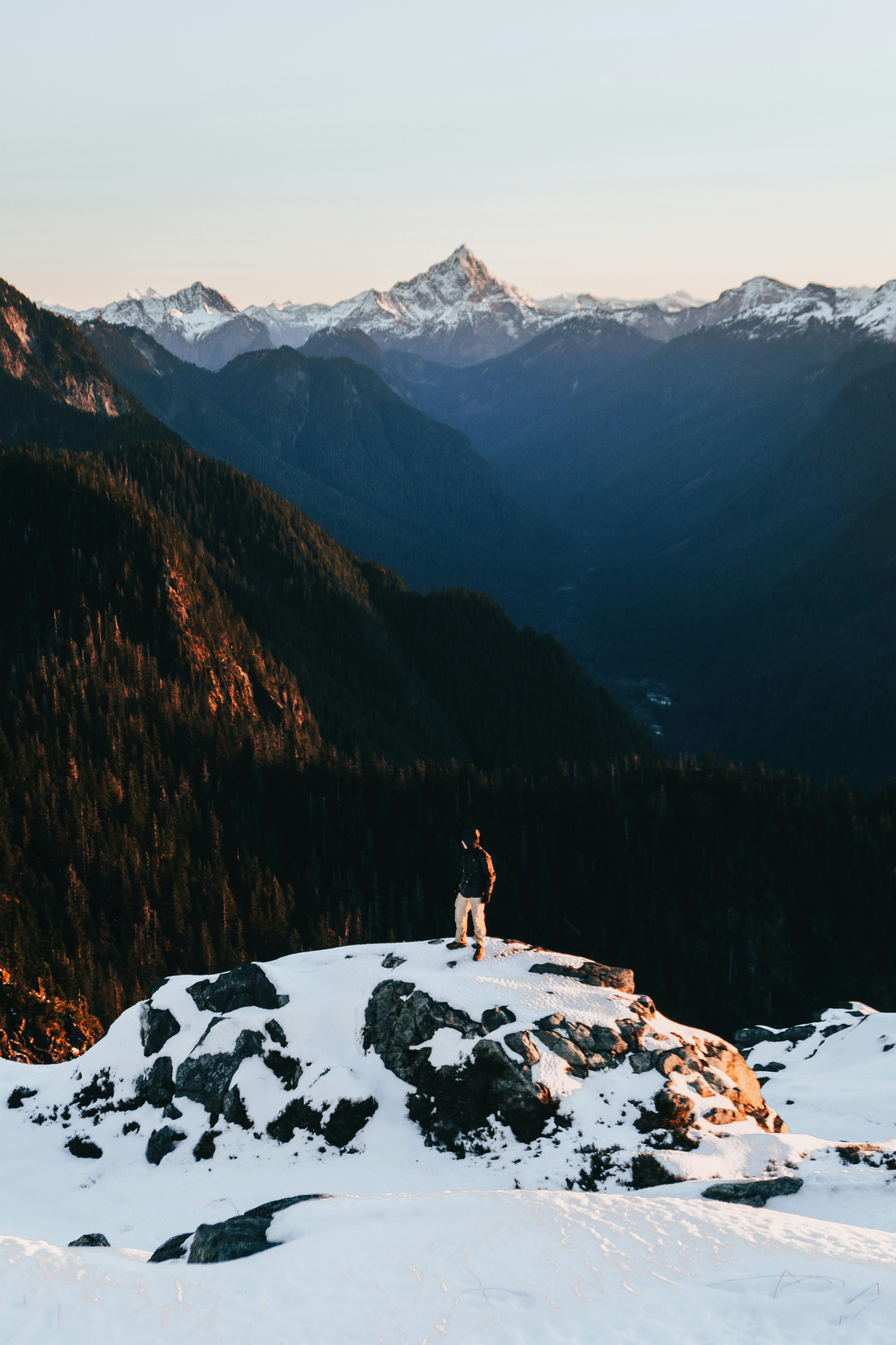 Individual standing atop a snowy peak, gazing at distant mountain ranges during sunset. The scene captures the tranquility of nature and the beauty of the wilderness.