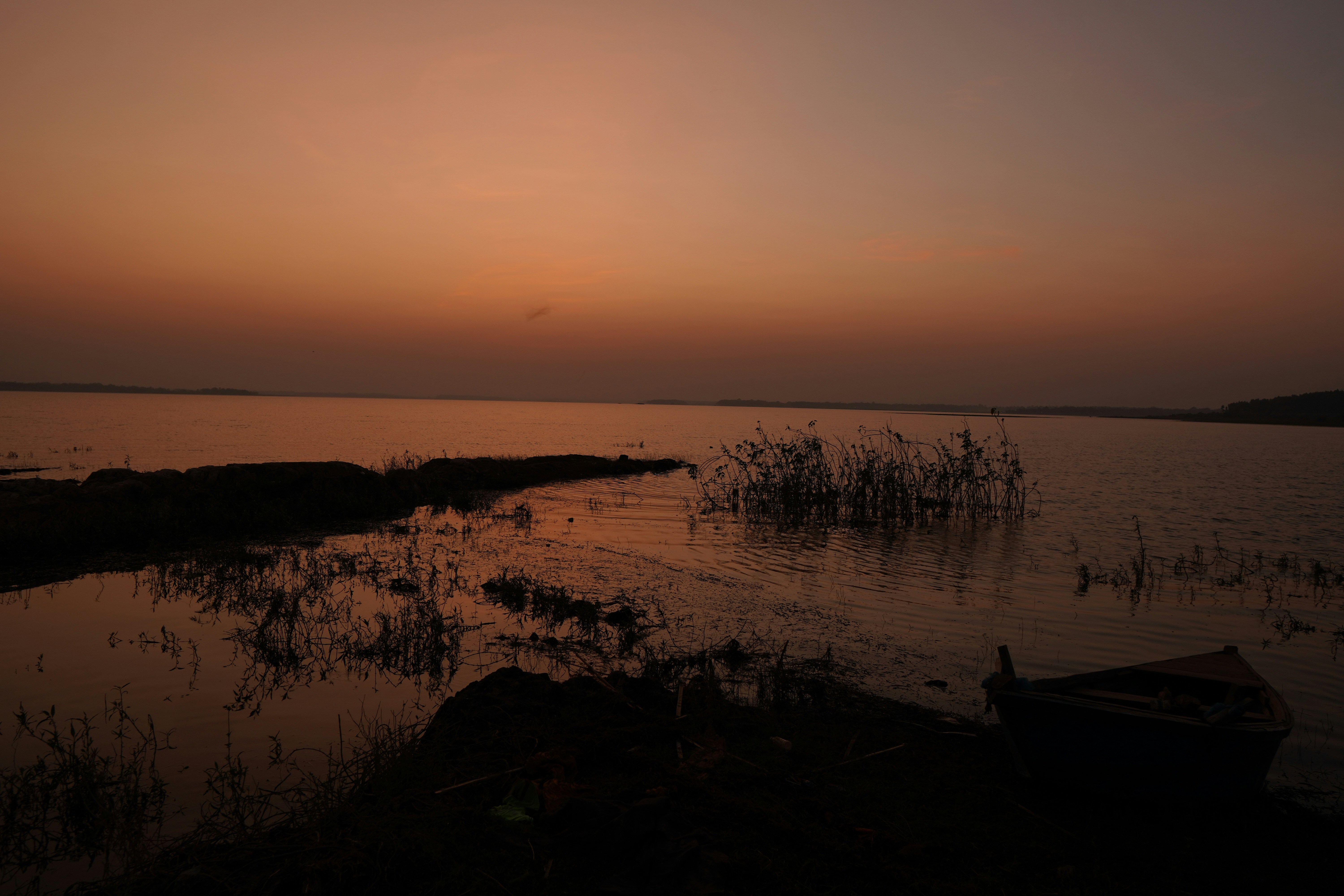 Serene lakeside at dusk, featuring silhouettes of vegetation and a solitary boat against a gradient sky. 