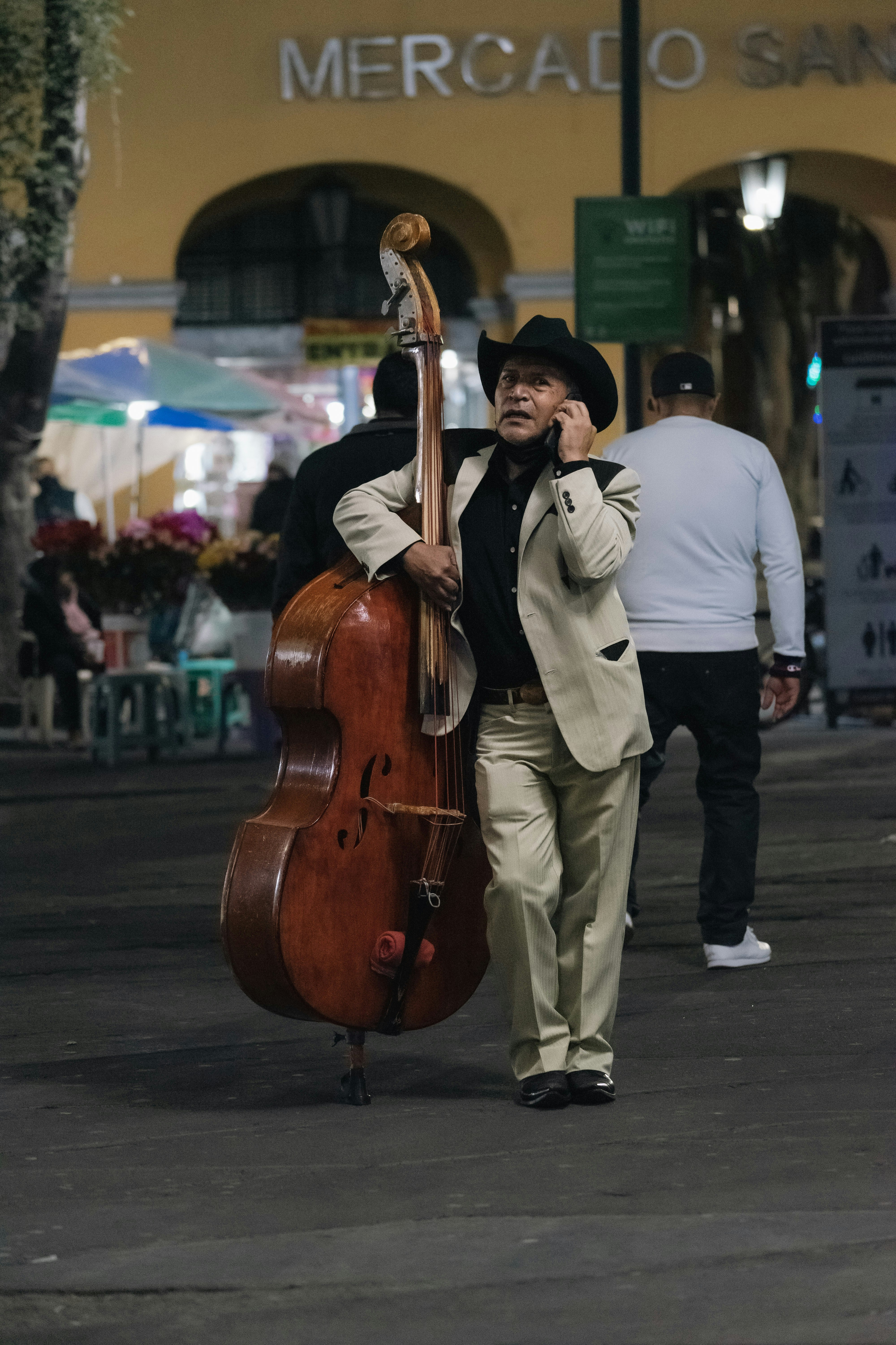 Musician in a cream suit converses on the phone while holding a double bass amidst a bustling market scene at night.