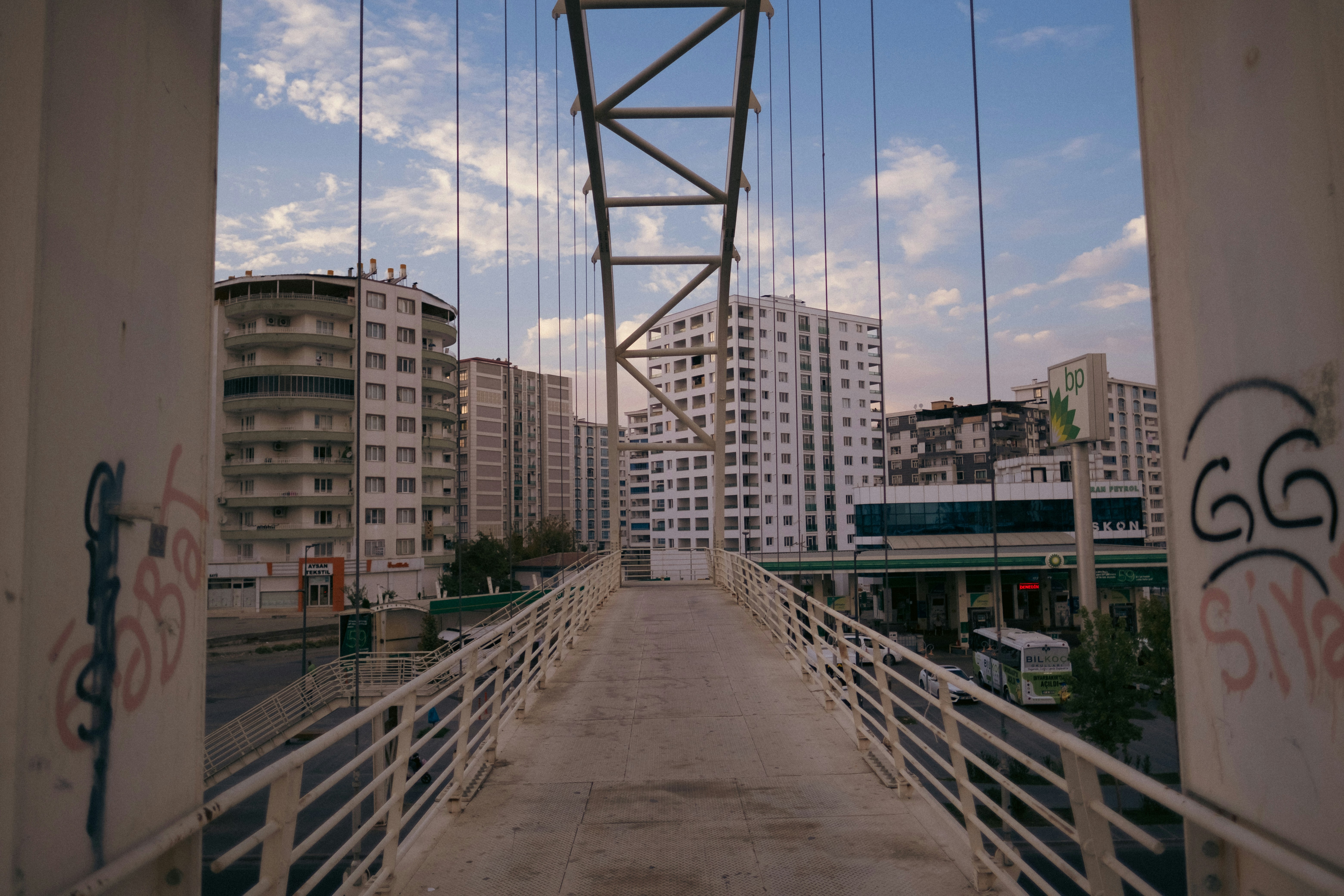 Suspended pedestrian bridge with urban graffiti, leading towards modern buildings under a cloudy sky.