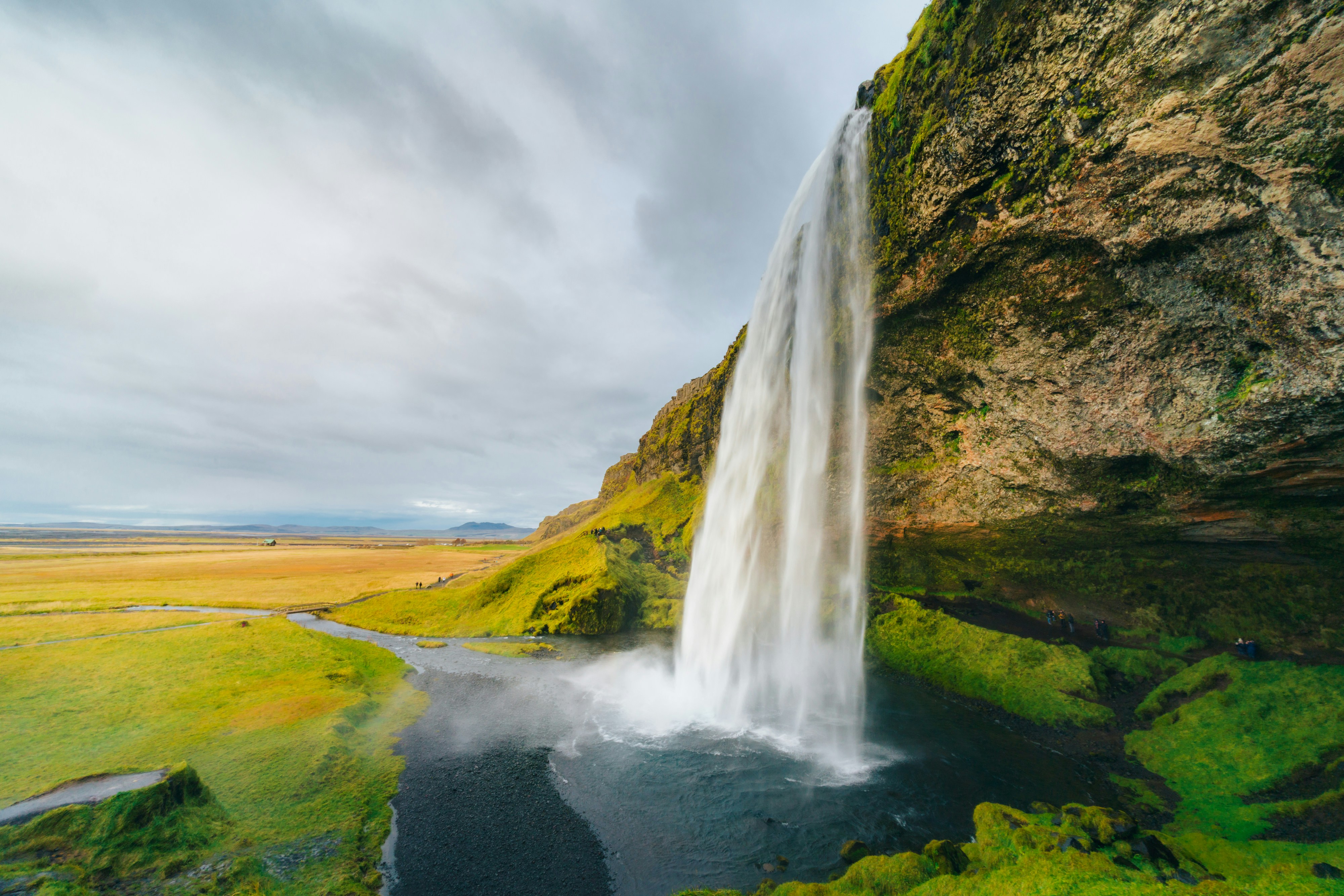 Majestic waterfall cascading over rugged cliffs, surrounded by lush greenery and open fields. The scene captures the tranquil beauty of Icelandic landscapes.
