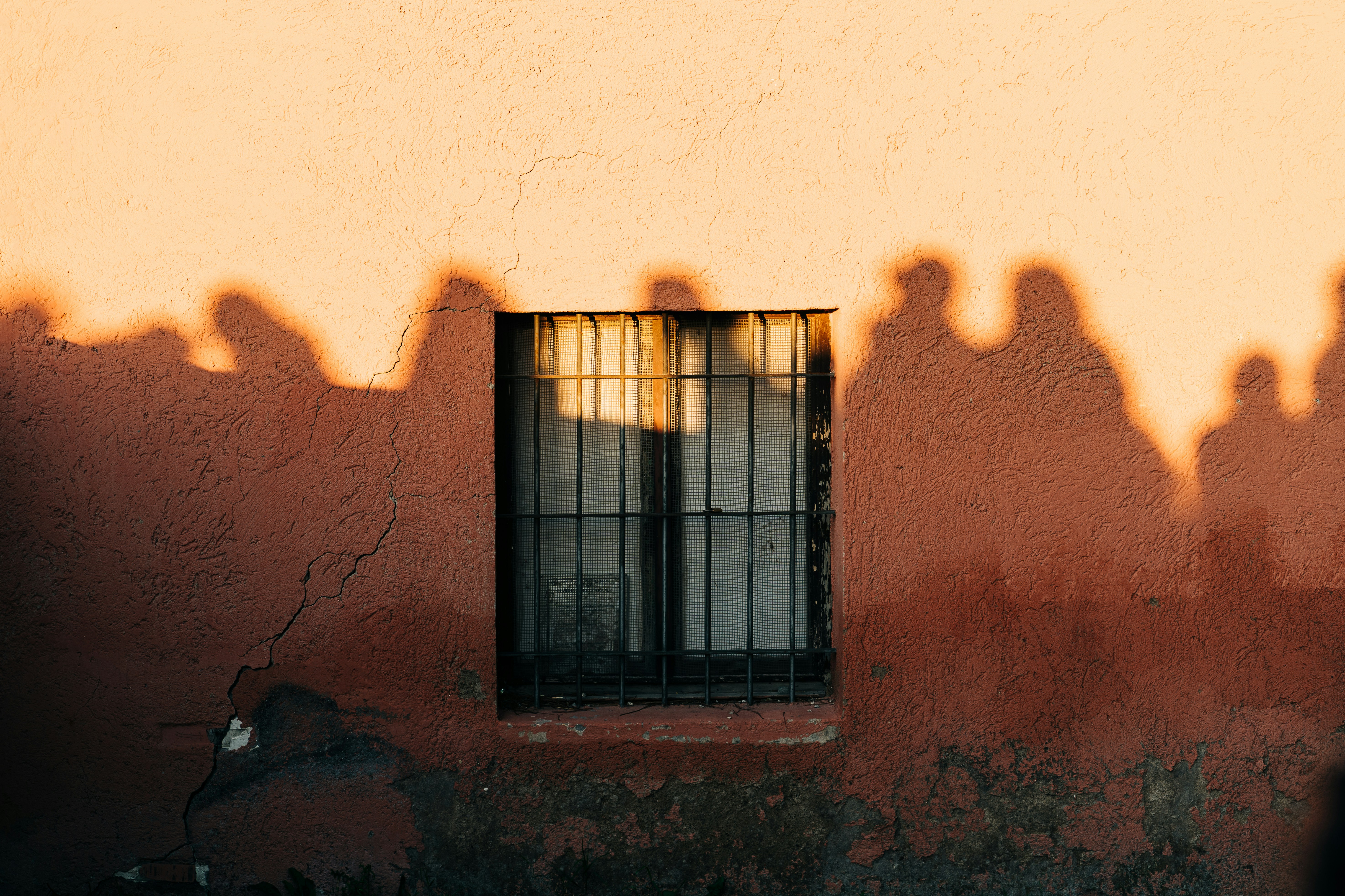 Shadows of figures dance across a textured orange wall, framing a barred window that hints at hidden stories behind it.