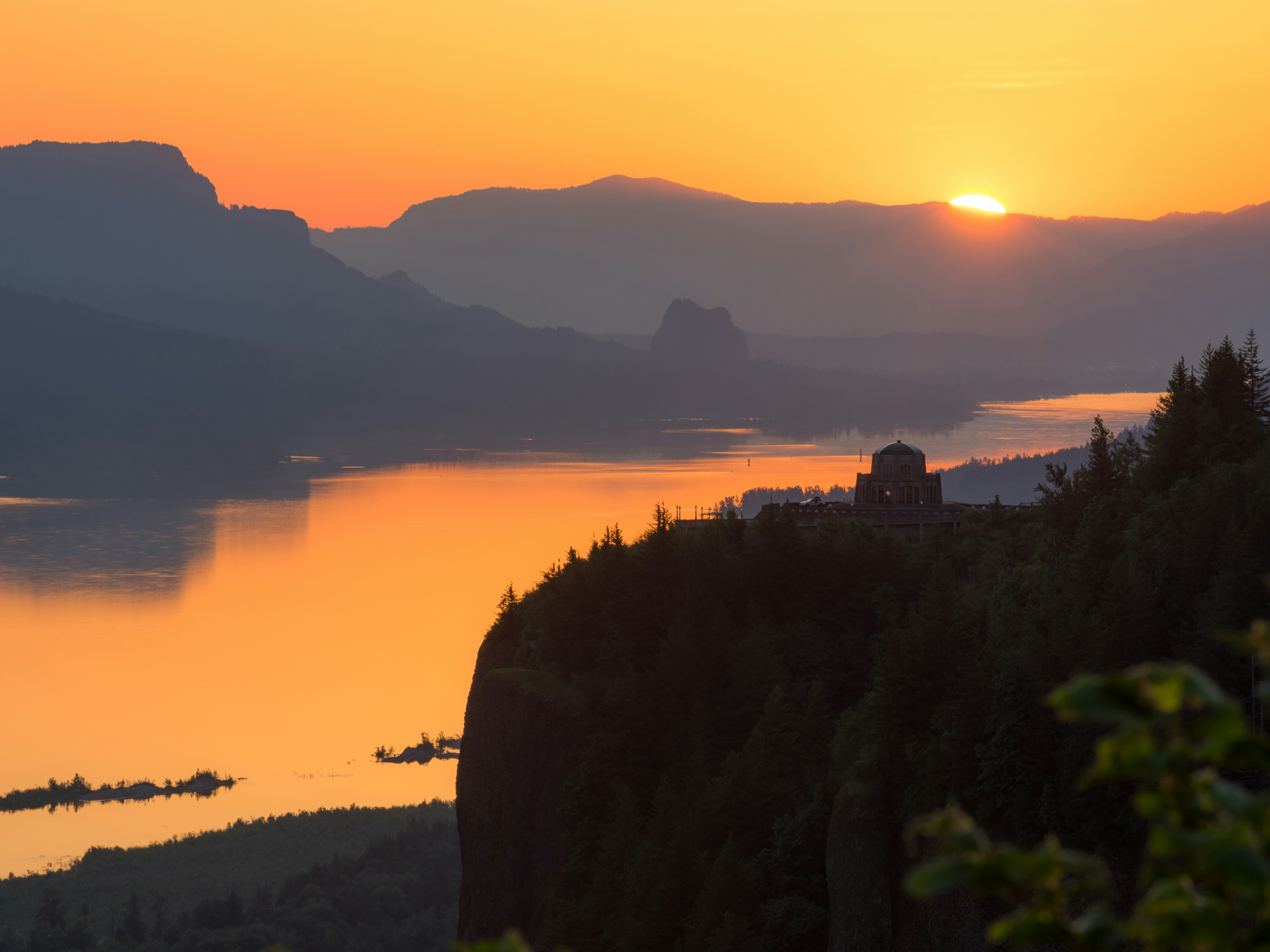 Golden sunset casting vibrant reflections over a tranquil river, framed by distant mountains and lush greenery.
