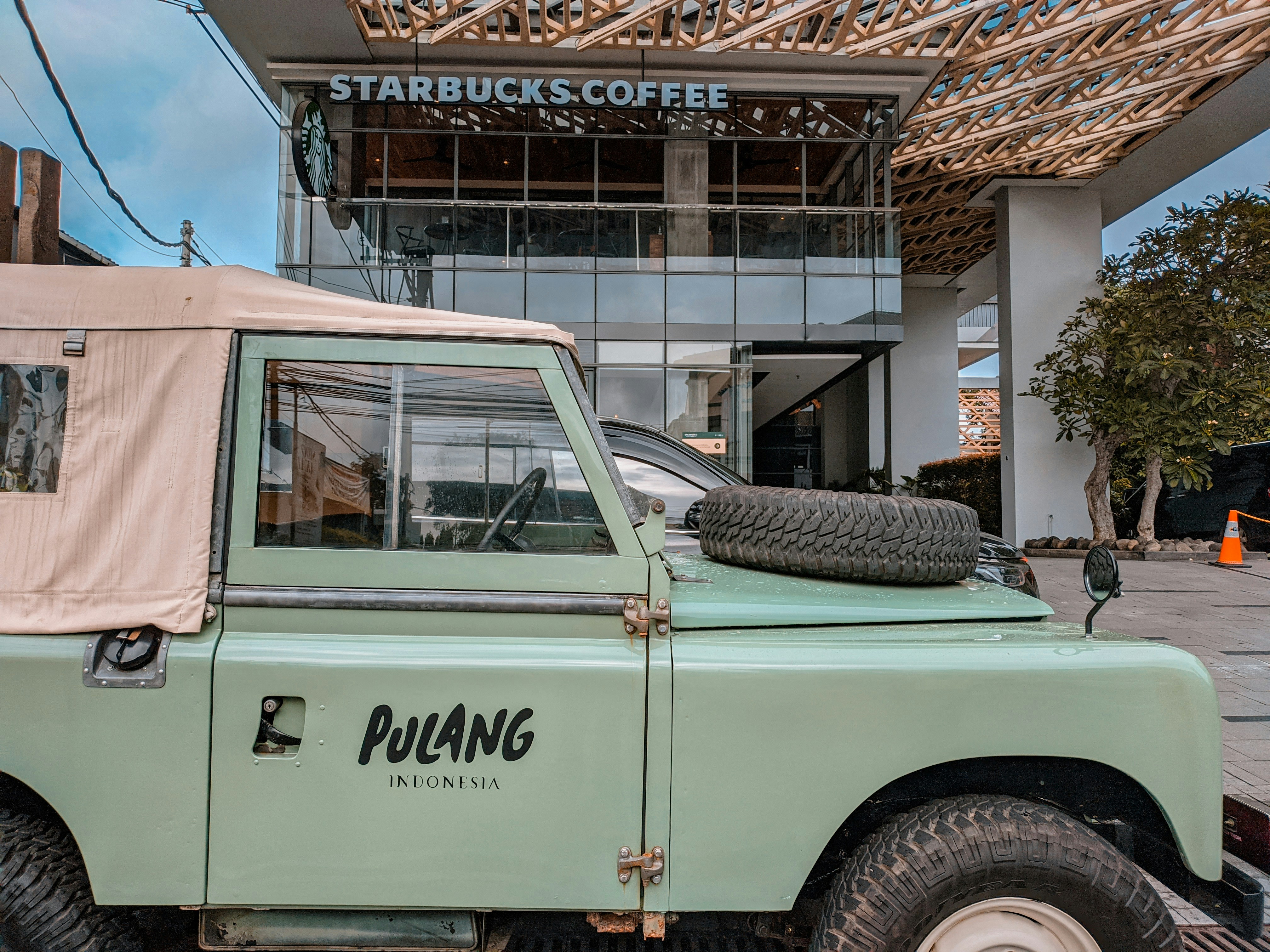 A classic green jeep parked in front of a contemporary Starbucks Coffee location, showcasing a blend of styles and cultures. The vehicle features the word 'Pulang' prominently displayed.