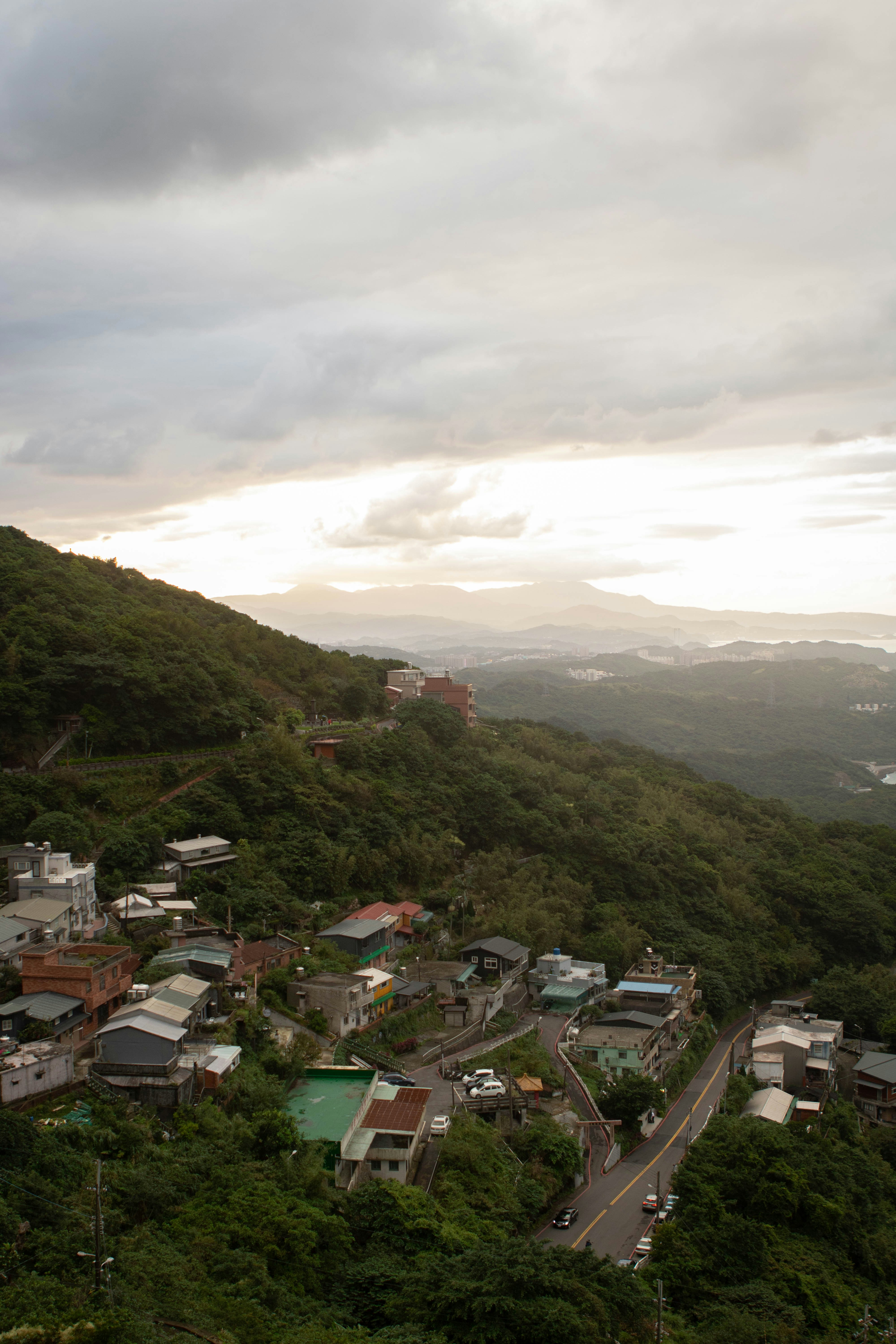 A panoramic view of a hillside village nestled among lush greenery, showcasing winding roads and quaint homes under a cloudy sky.