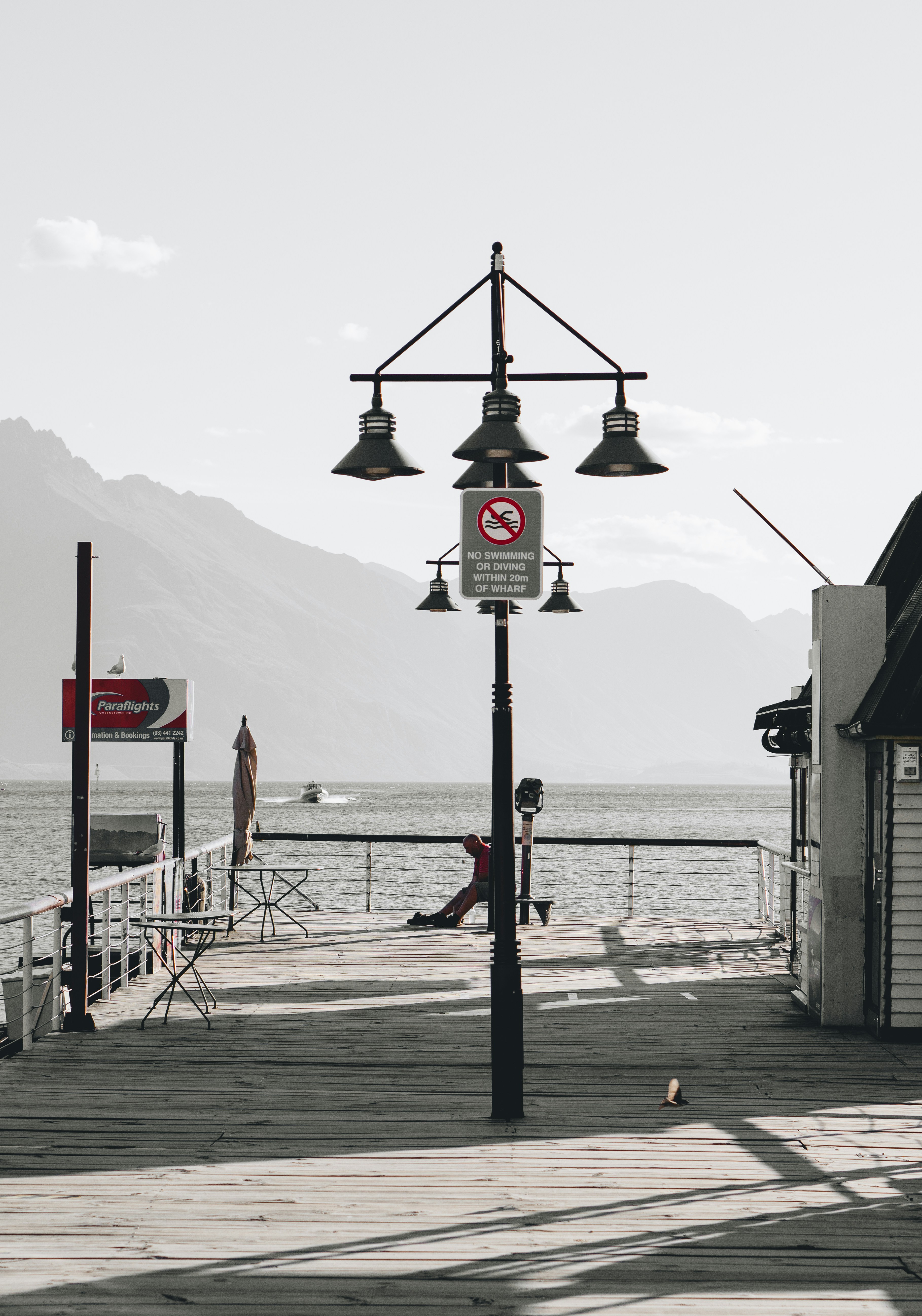 A serene pier scene featuring hanging lights and a no swimming sign, with a distant boat gliding across the water. The composition highlights the interplay of light and shadow.