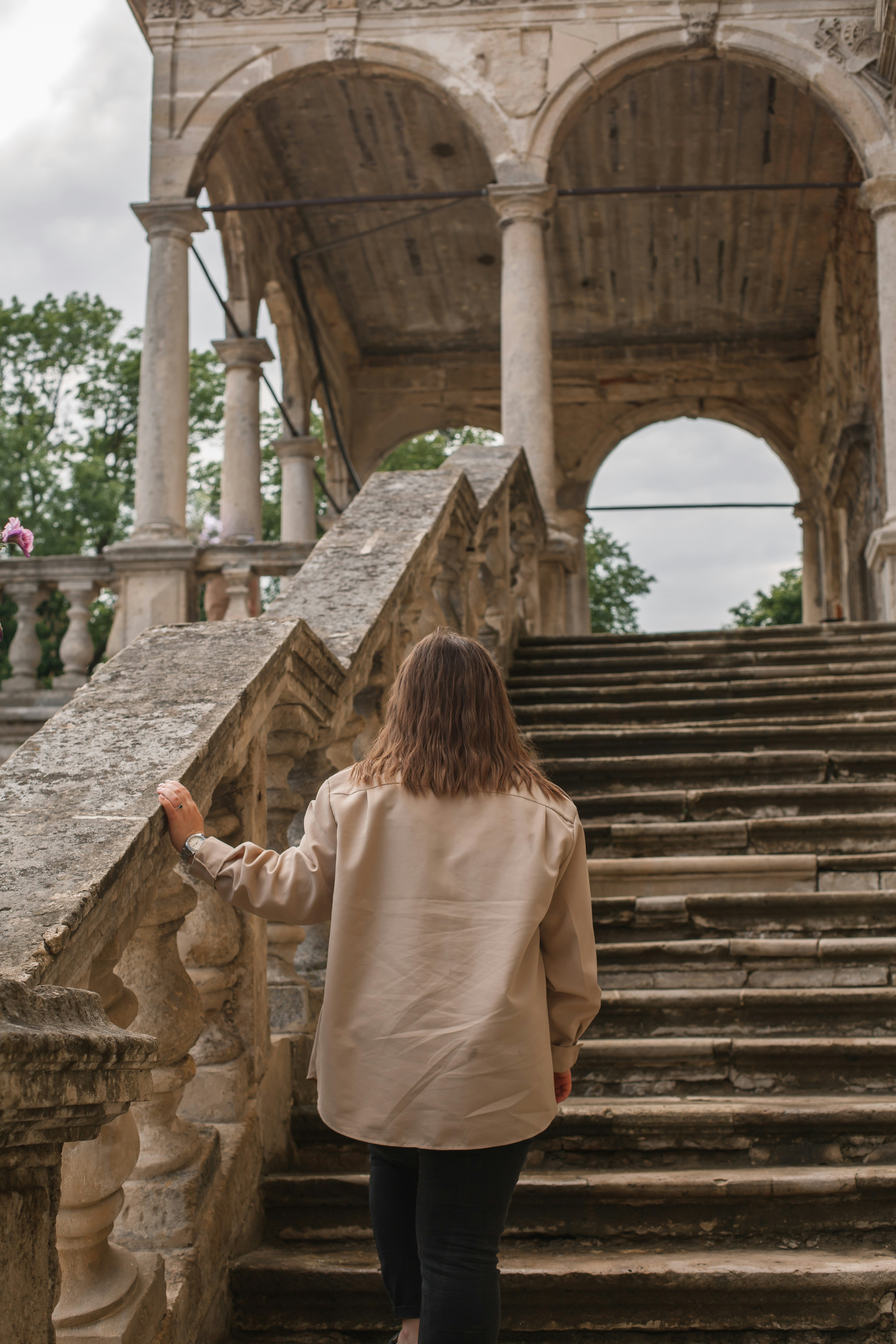 A person in a light beige shirt ascends a weathered staircase, flanked by ornate railings and classical architecture, leading to an arched passageway. The scene evokes a sense of exploration and history.