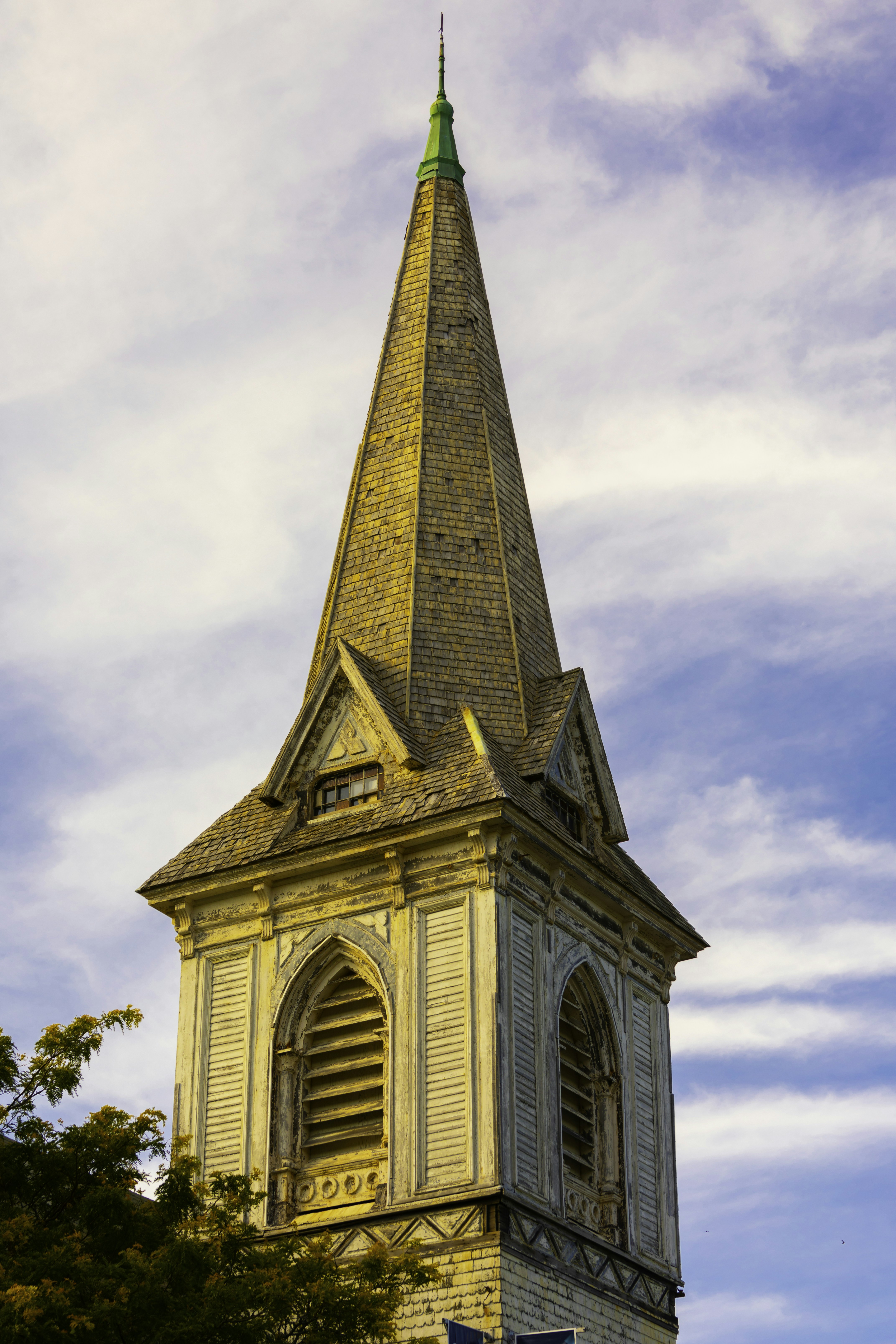 Weathered church steeple with intricate wooden details and a green-tipped spire against a cloudy sky.
