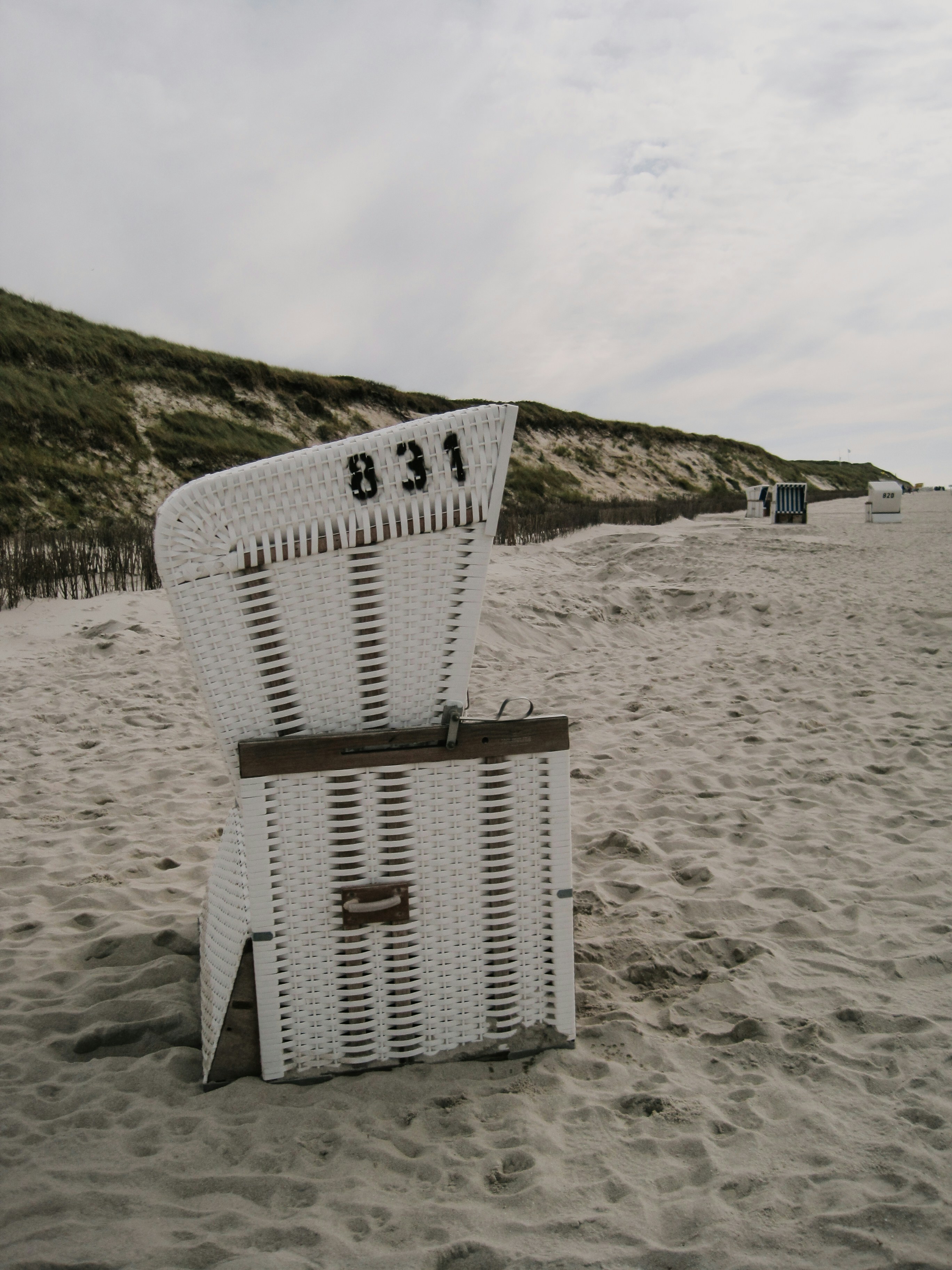 Weathered beach chair numbered 831 stands in soft sand, framed by gentle dunes and a cloudy sky.